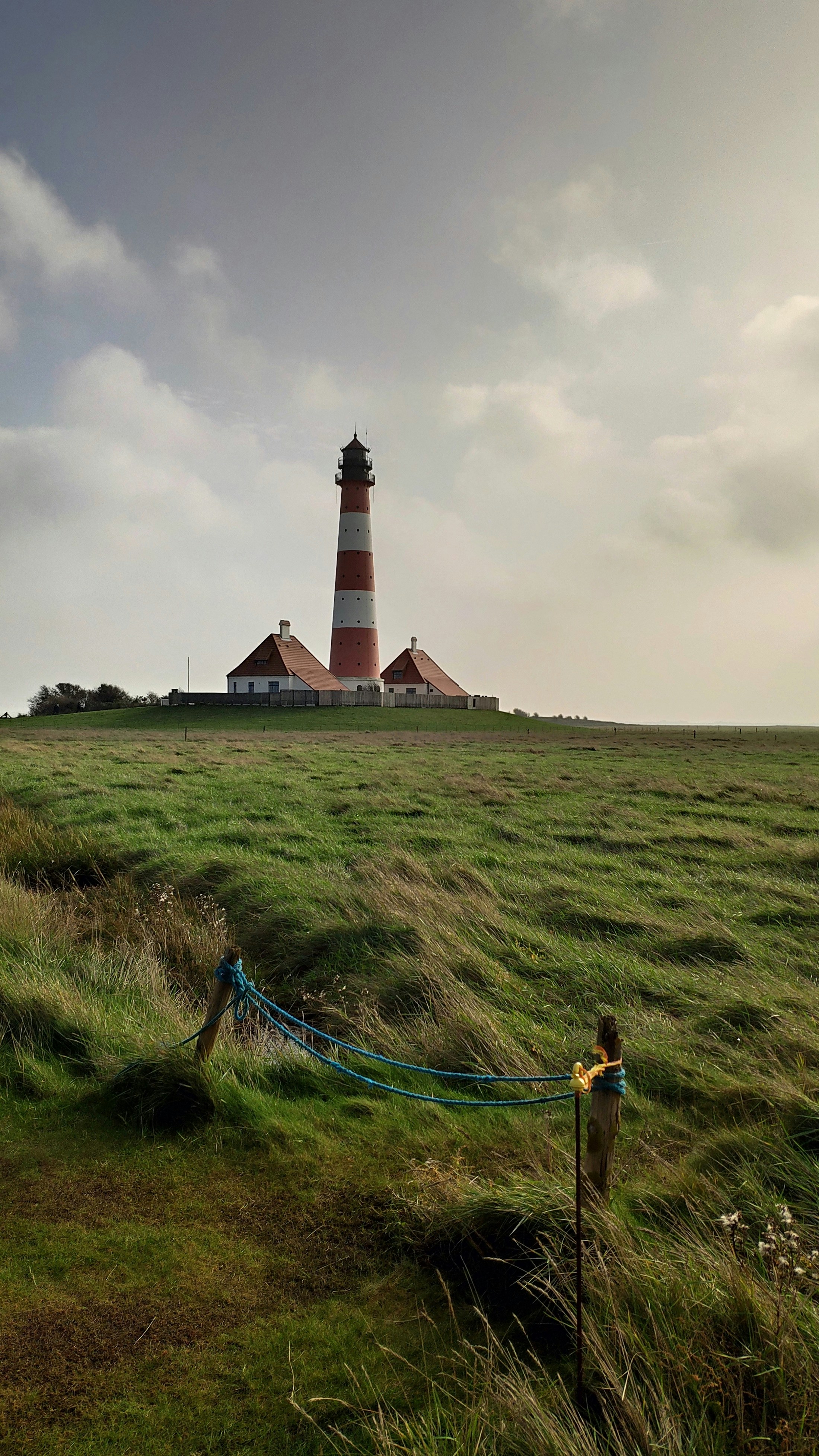 A photograph of a red-and-white striped lighthouse atop a grassy hill, with a blue rope barrier and a person in the foreground.
