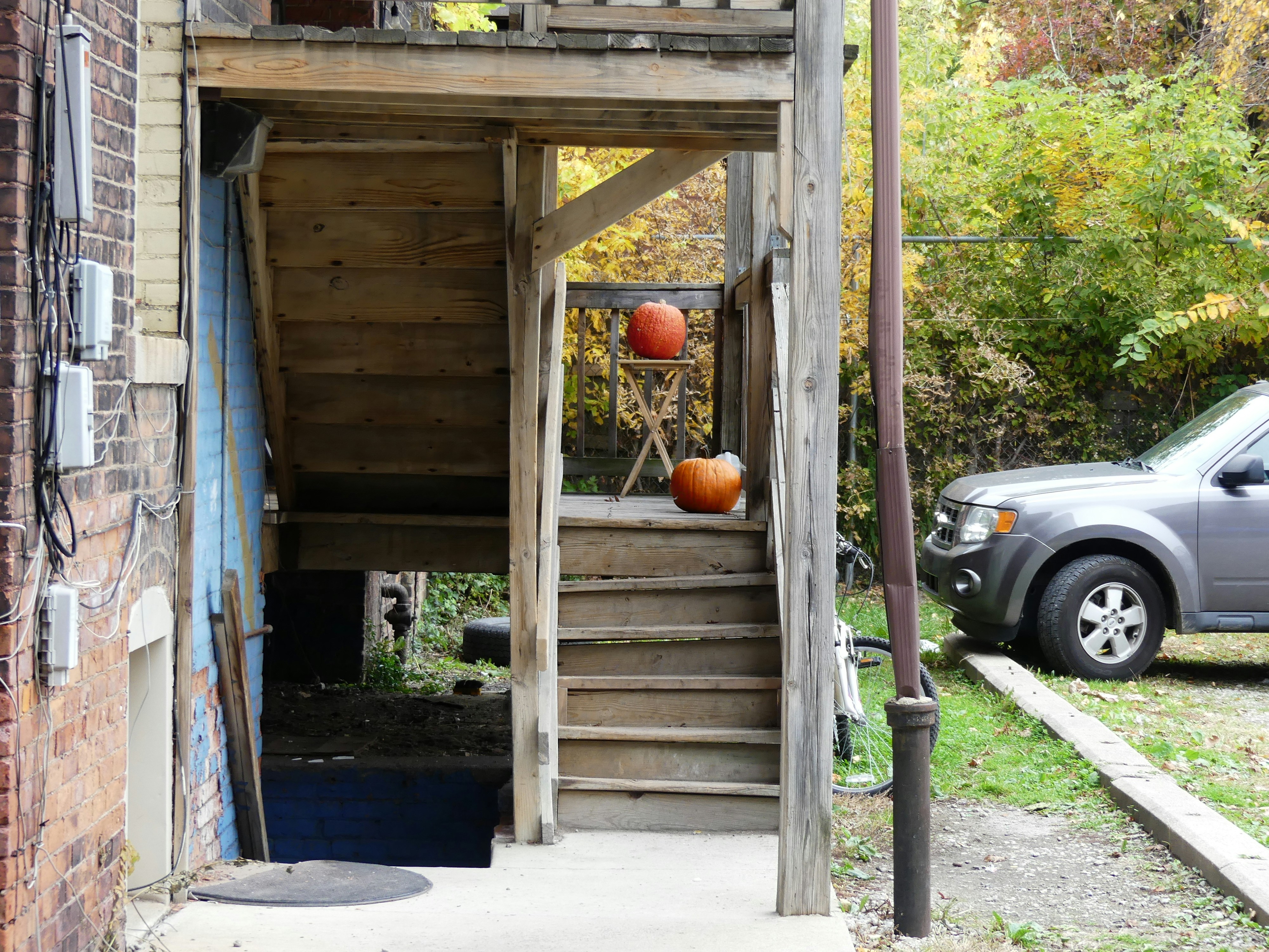 Wooden stairs leading to a shaded area adorned with pumpkins, surrounded by vibrant foliage. A parked vehicle is partially visible in the background.