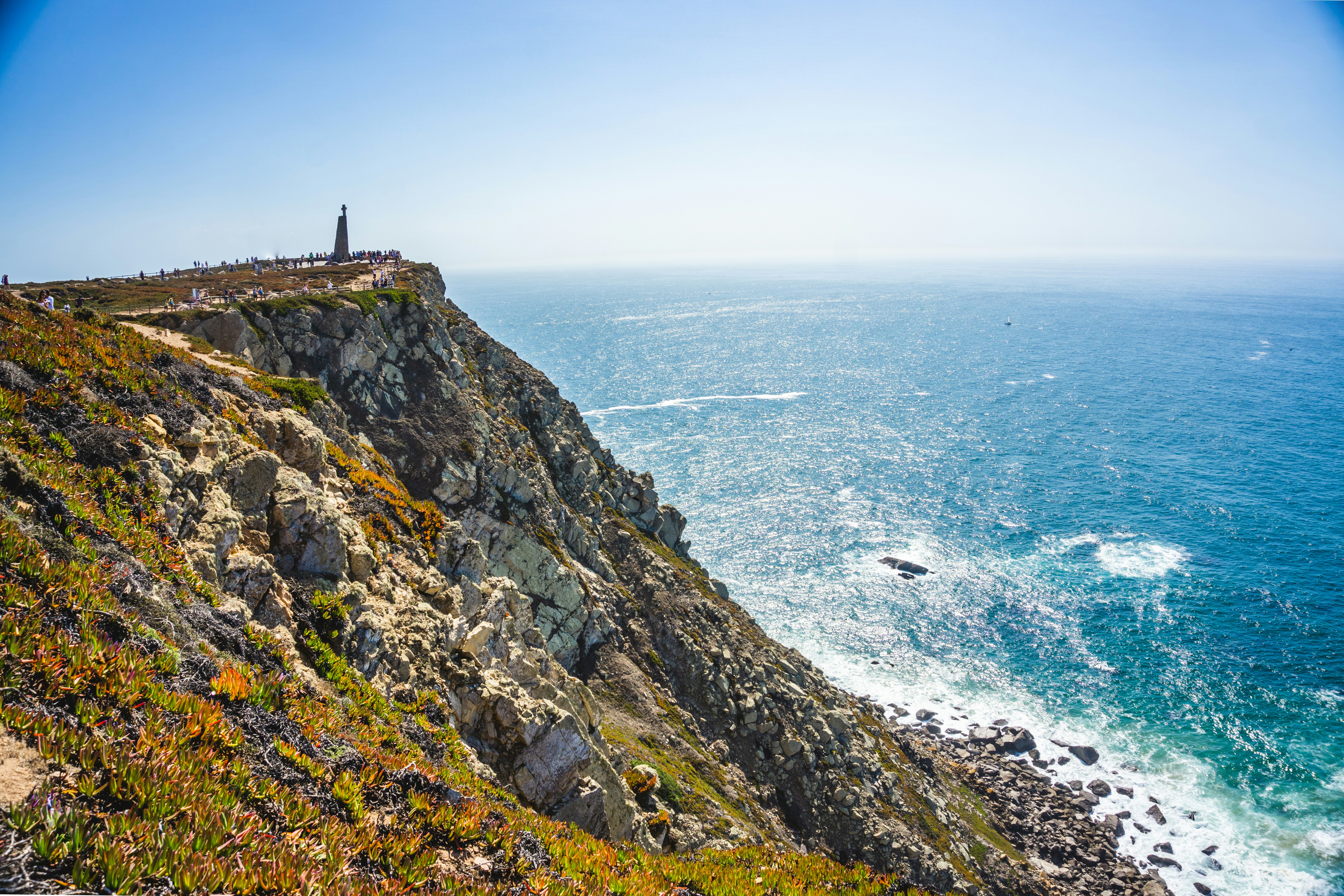 A lighthouse stands proudly atop a rugged cliff, gazing out over the vibrant blue ocean waves crashing against the rocky shore.
