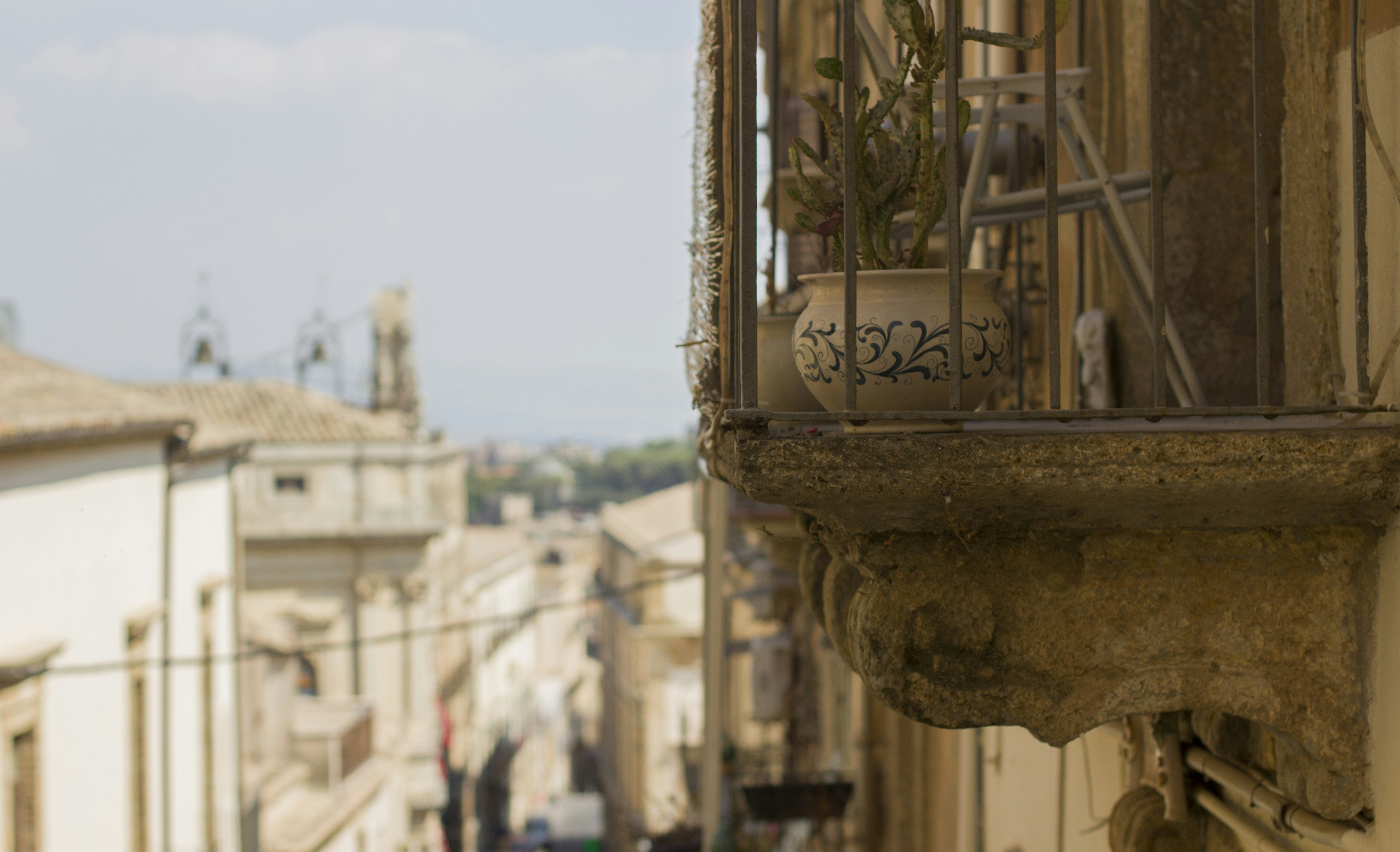 A charming balcony adorned with a potted plant overlooks a narrow street, revealing a glimpse of historical architecture and a distant landscape.