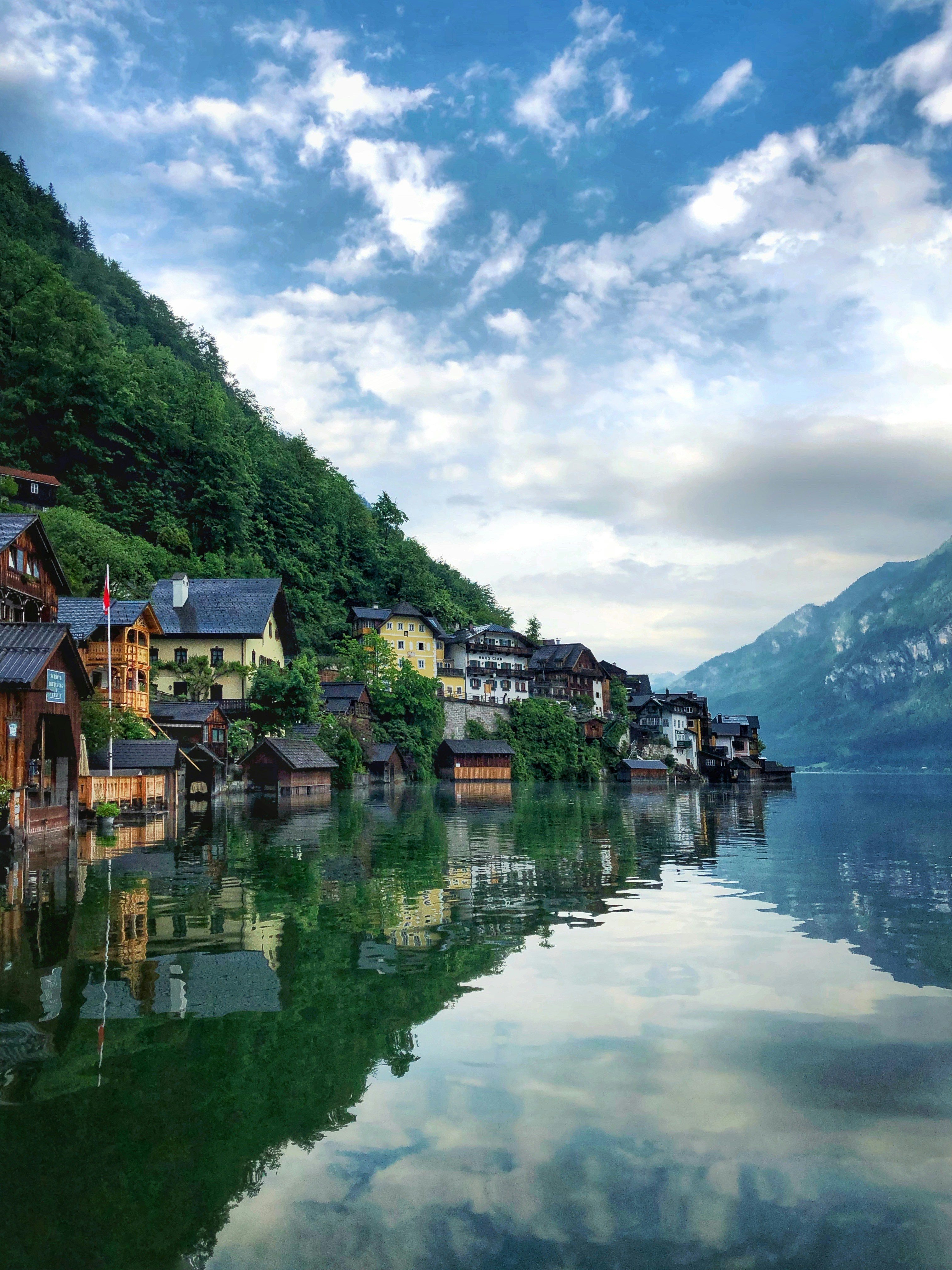 Charming lakeside village nestled against a lush mountainside, with colorful houses reflecting in tranquil waters under a partially cloudy sky.