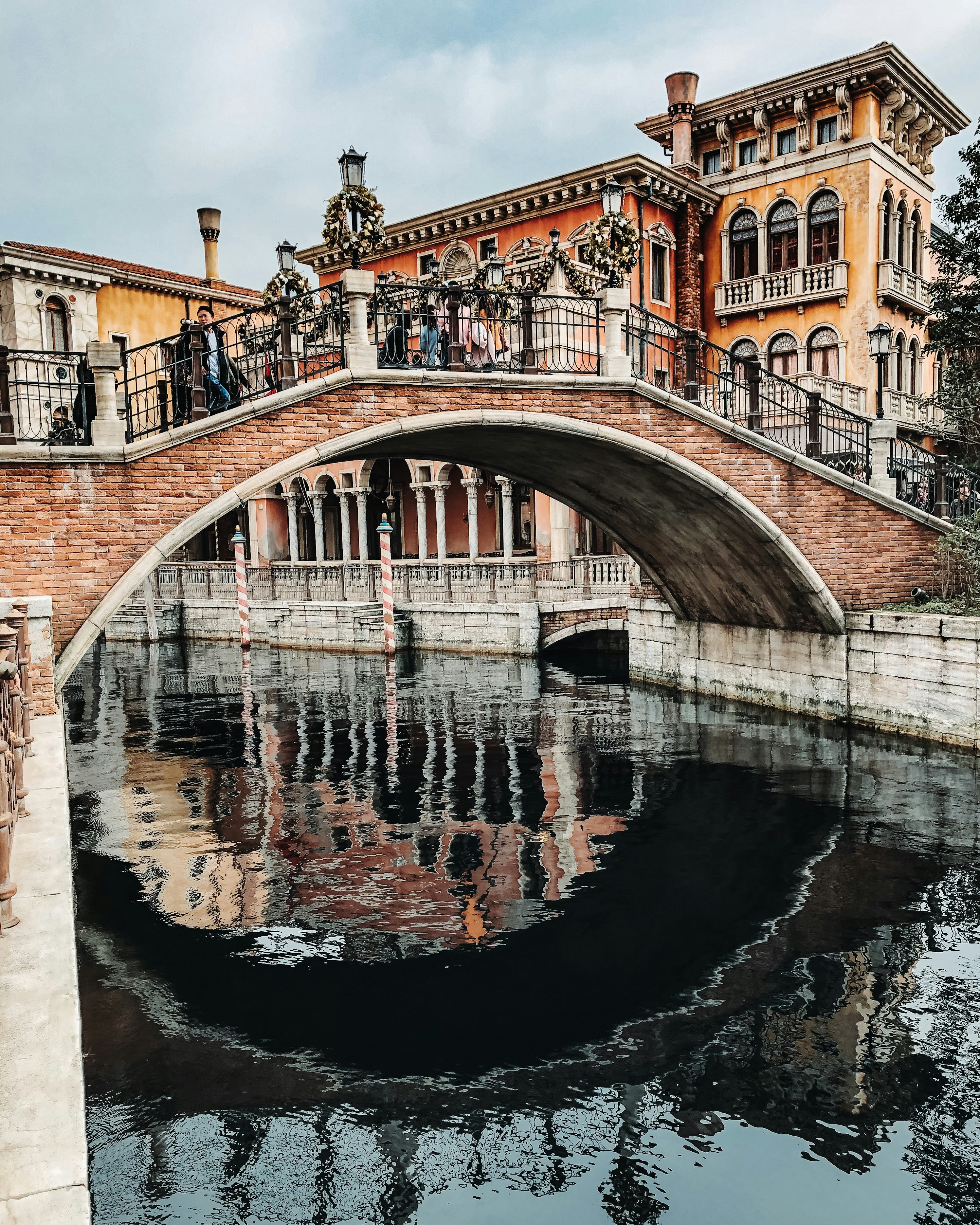 Arched bridge and ornate building reflecting in a calm canal under a cloudy sky.
