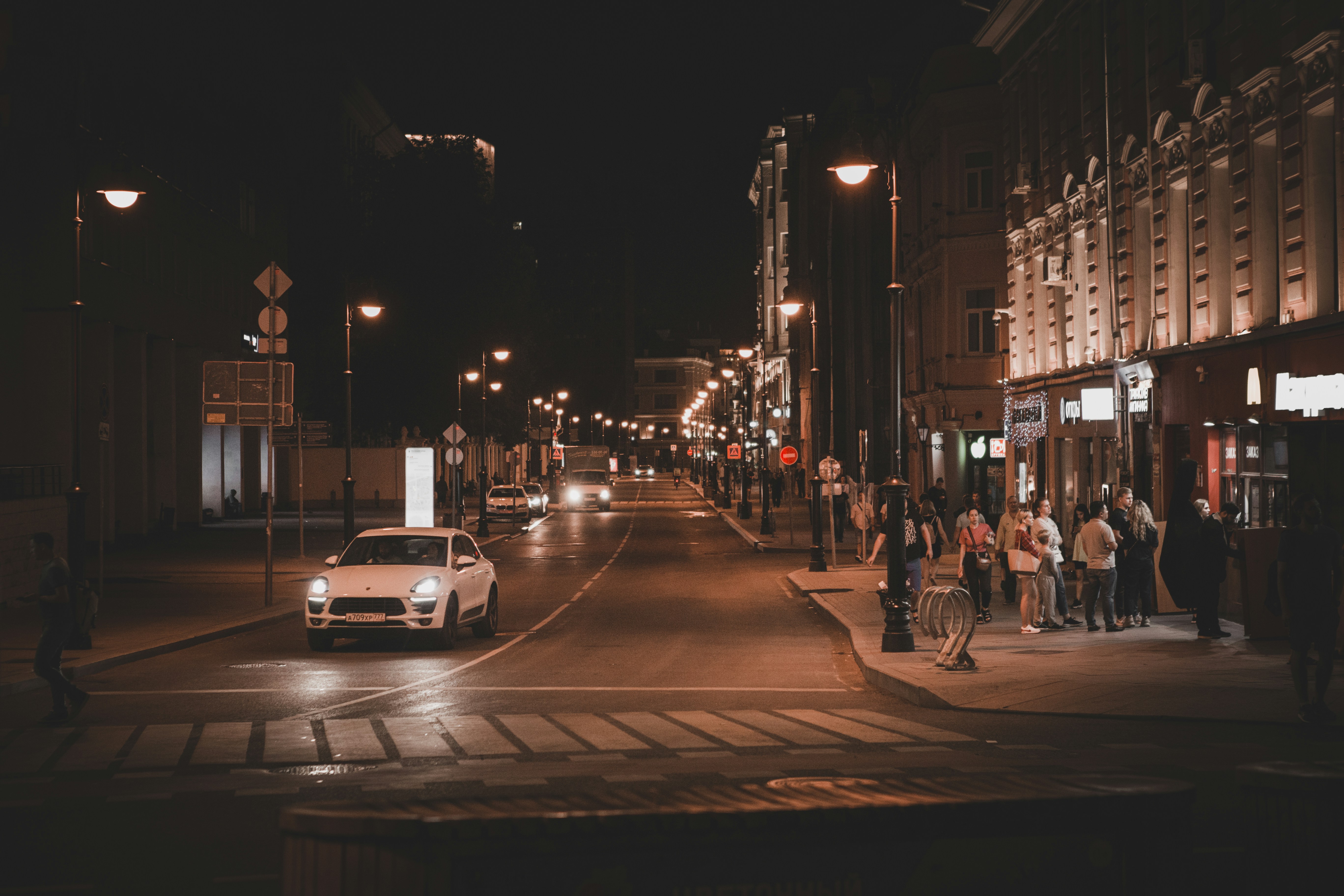 A sleek car navigates a quiet urban street illuminated by streetlights, while a group of pedestrians gathers nearby, showcasing the vibrant nightlife.