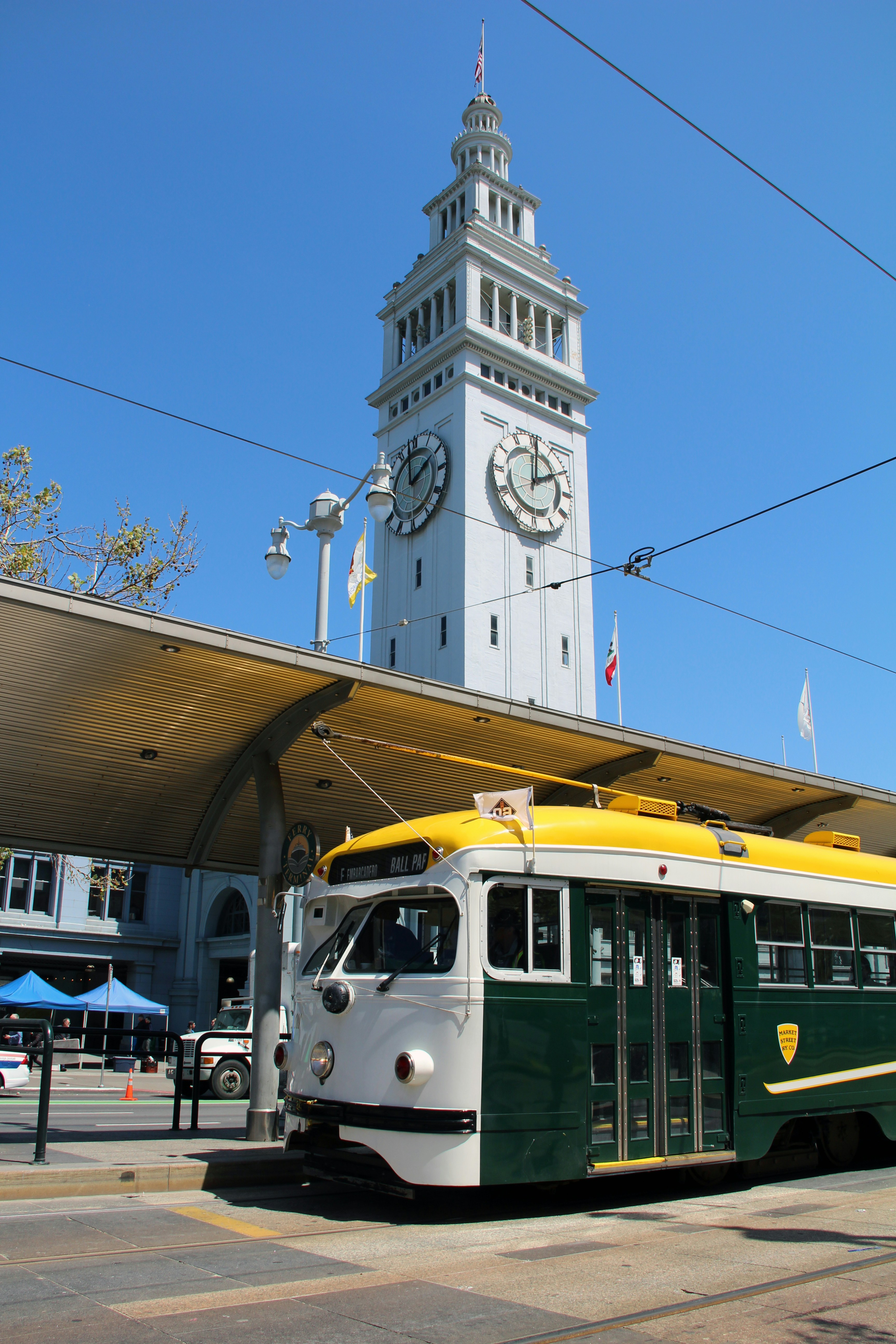 Classic green and yellow streetcar parked at a historic transit station with a prominent clock tower in the background. The scene captures a blend of transportation history and architectural elegance.