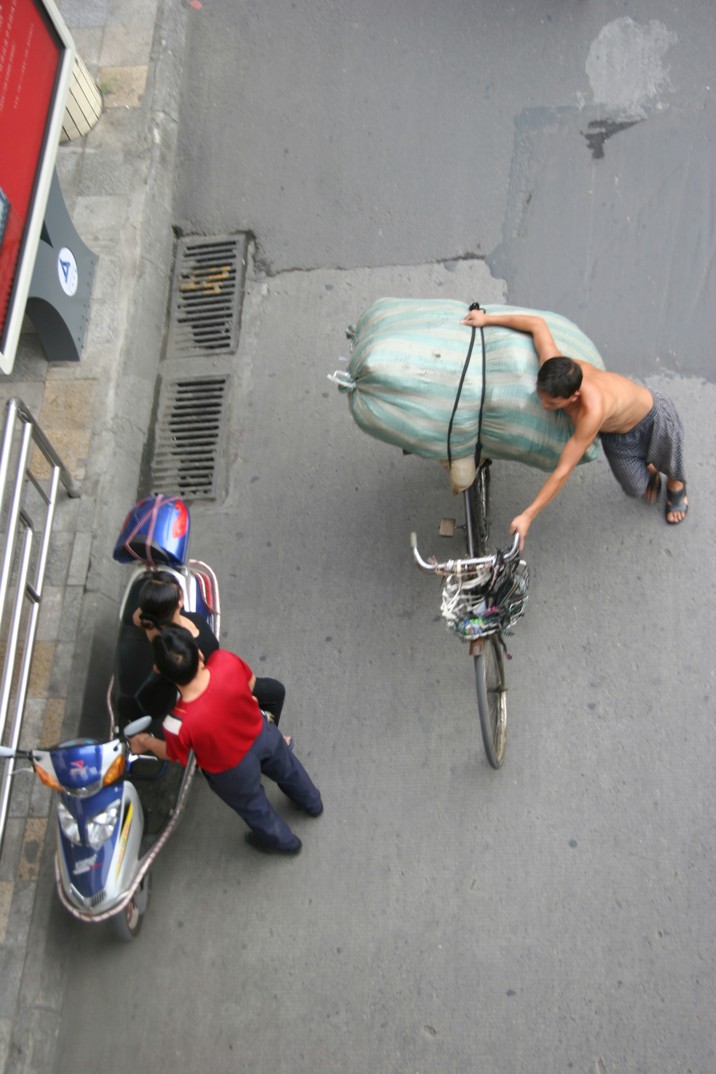 A shirtless man maneuvers a heavily loaded bicycle while two individuals converse nearby, capturing the essence of urban life and resilience.