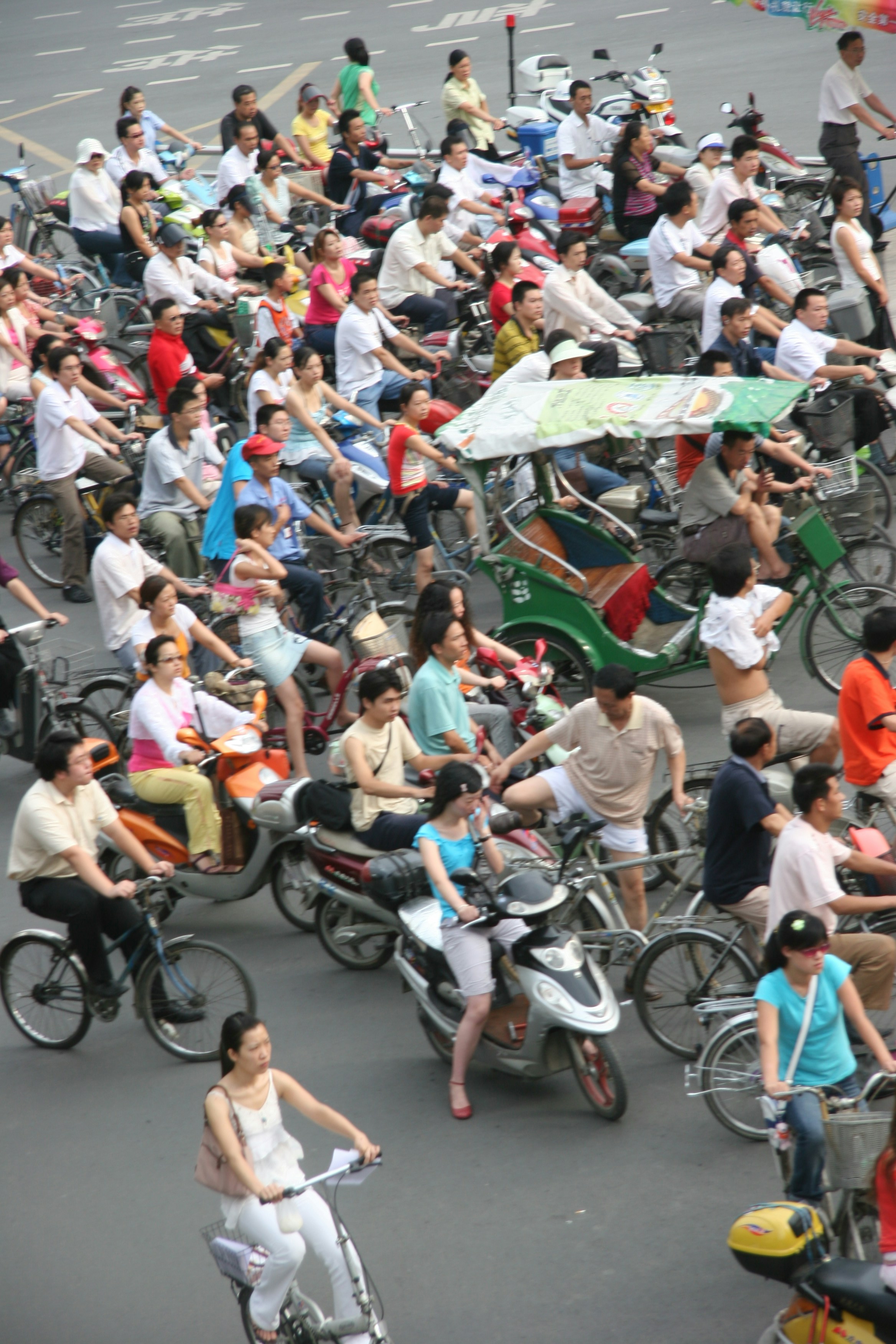 Dense city street scene crowded with pedestrians riding bicycles and motor scooters at a busy intersection. A colorful array of vehicles, including a green tricycle taxi, adds kinetic chaos.