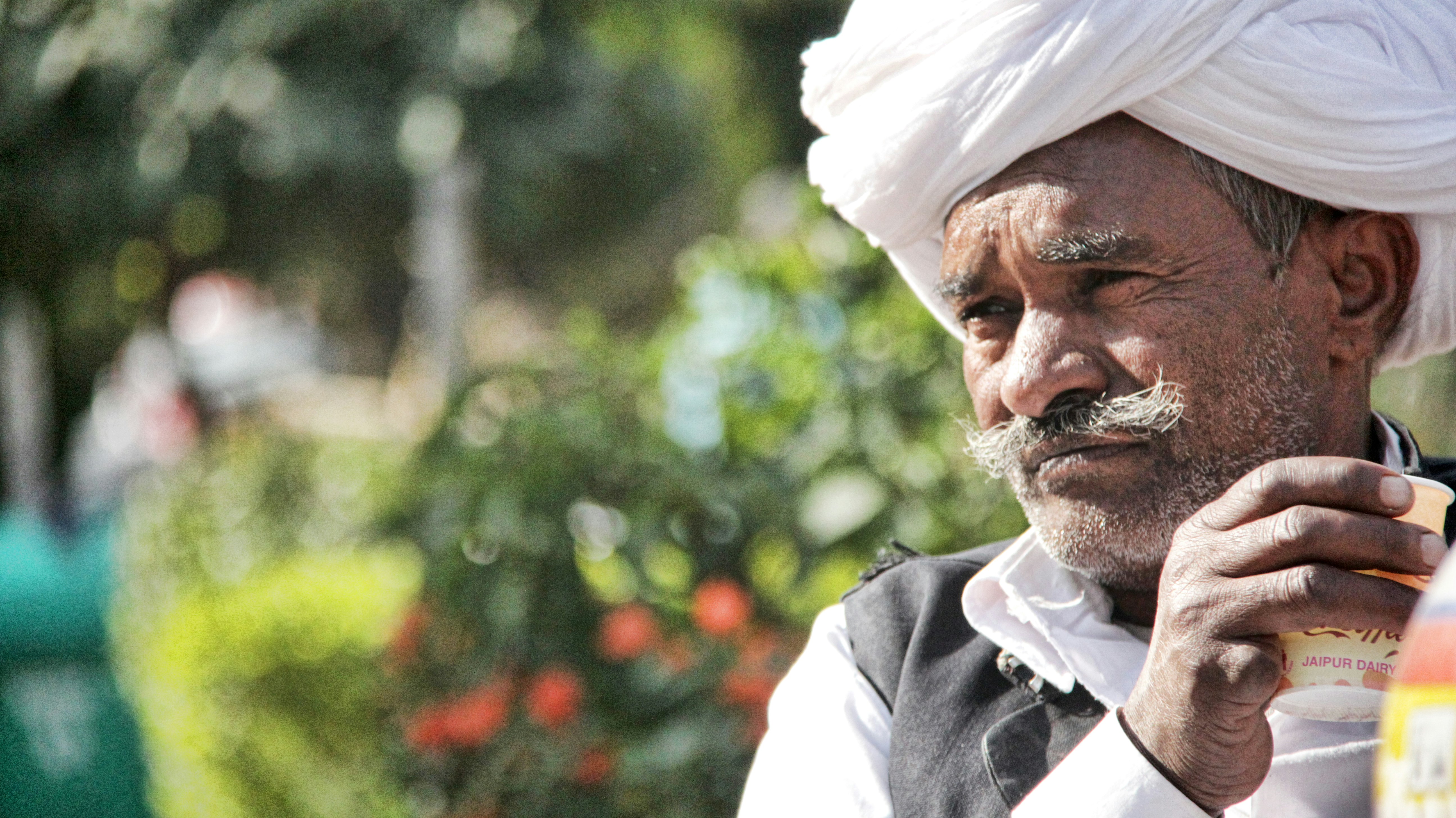 Elderly man in a white turban enjoying a drink outdoors with vibrant foliage in the background.
