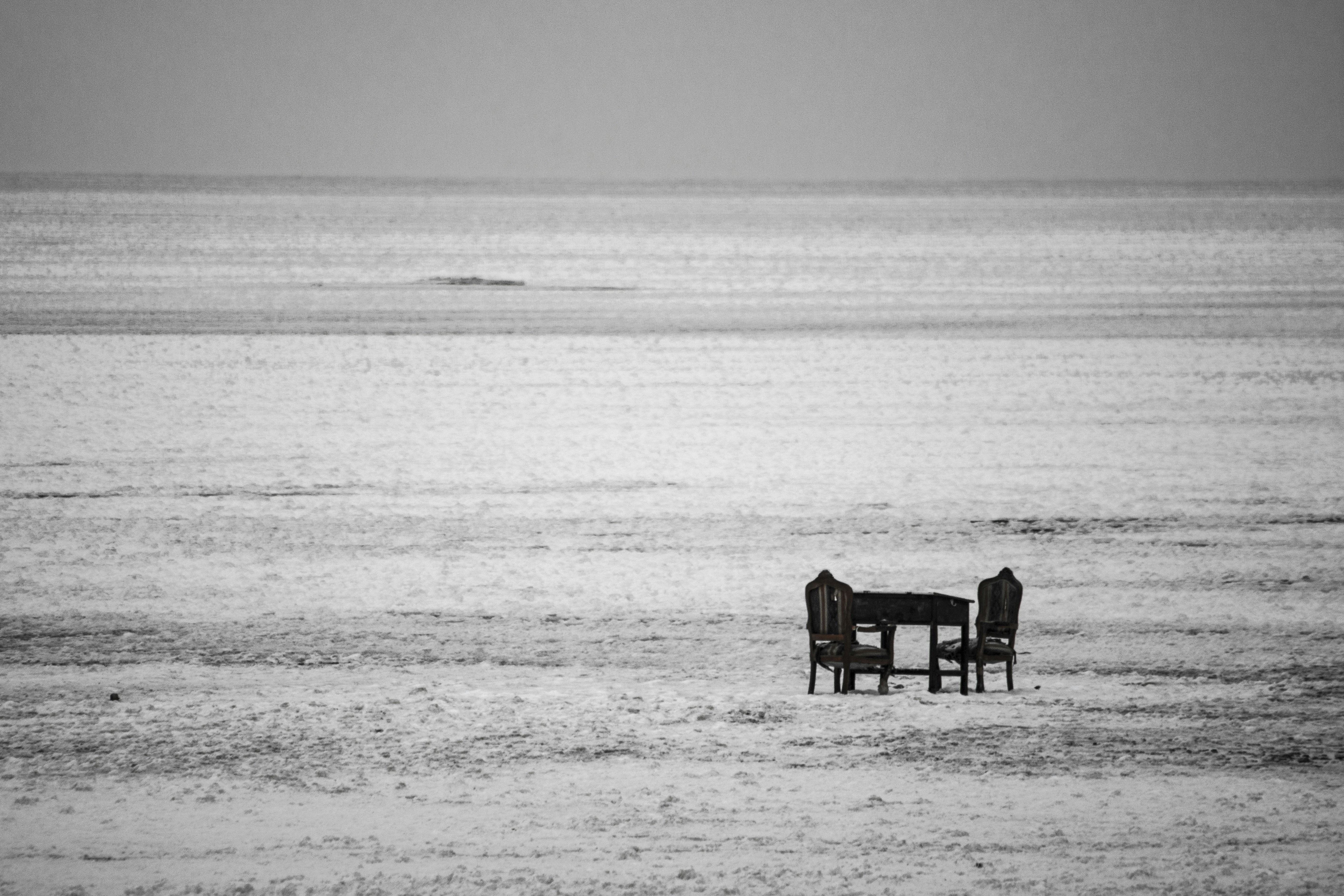 A table and two chairs stand alone on a vast, desolate shoreline, surrounded by a calm sea under a muted sky.