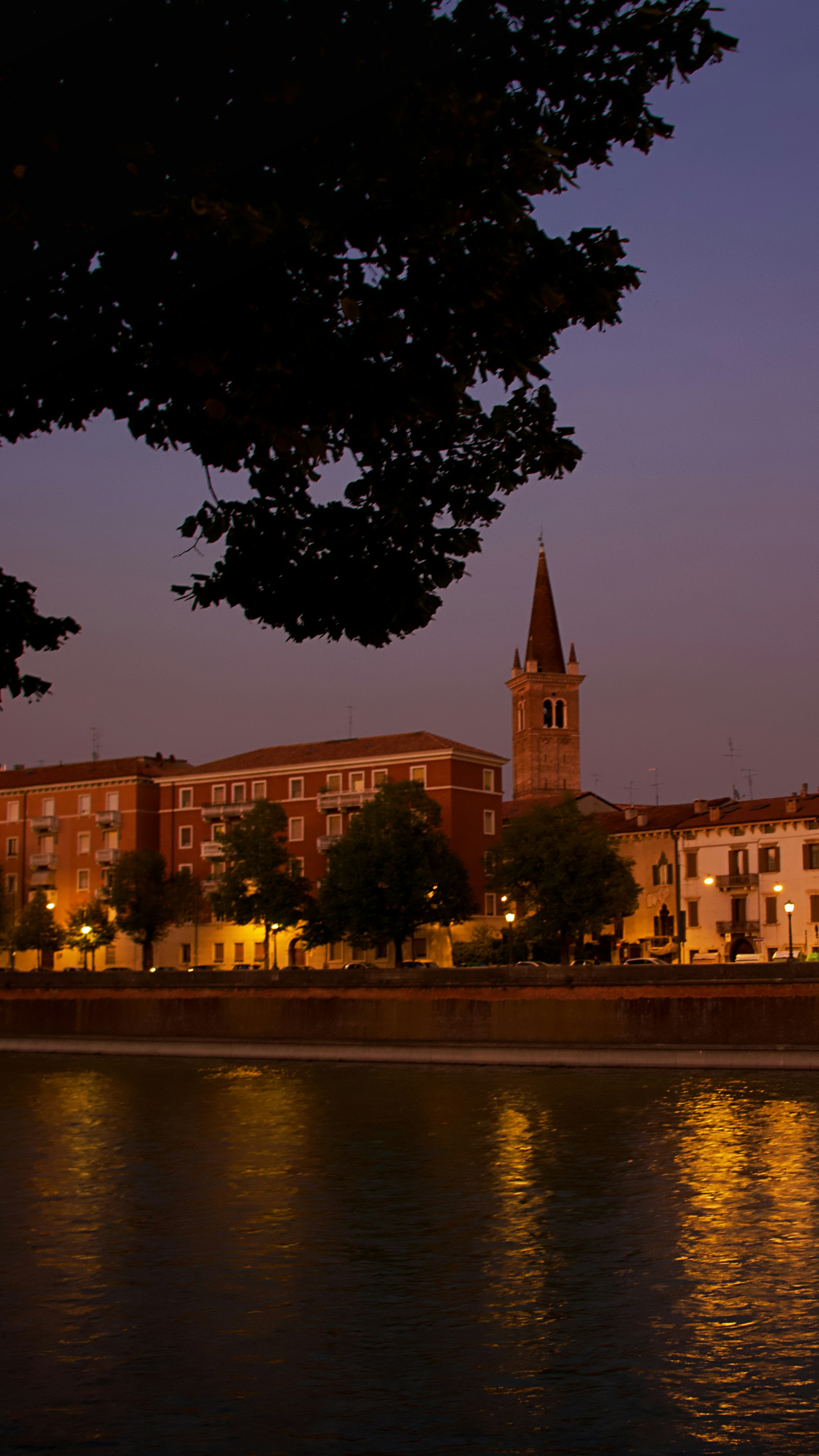 Historic buildings lining a tranquil river at twilight, with a prominent clock tower and soft reflections on the water's surface.