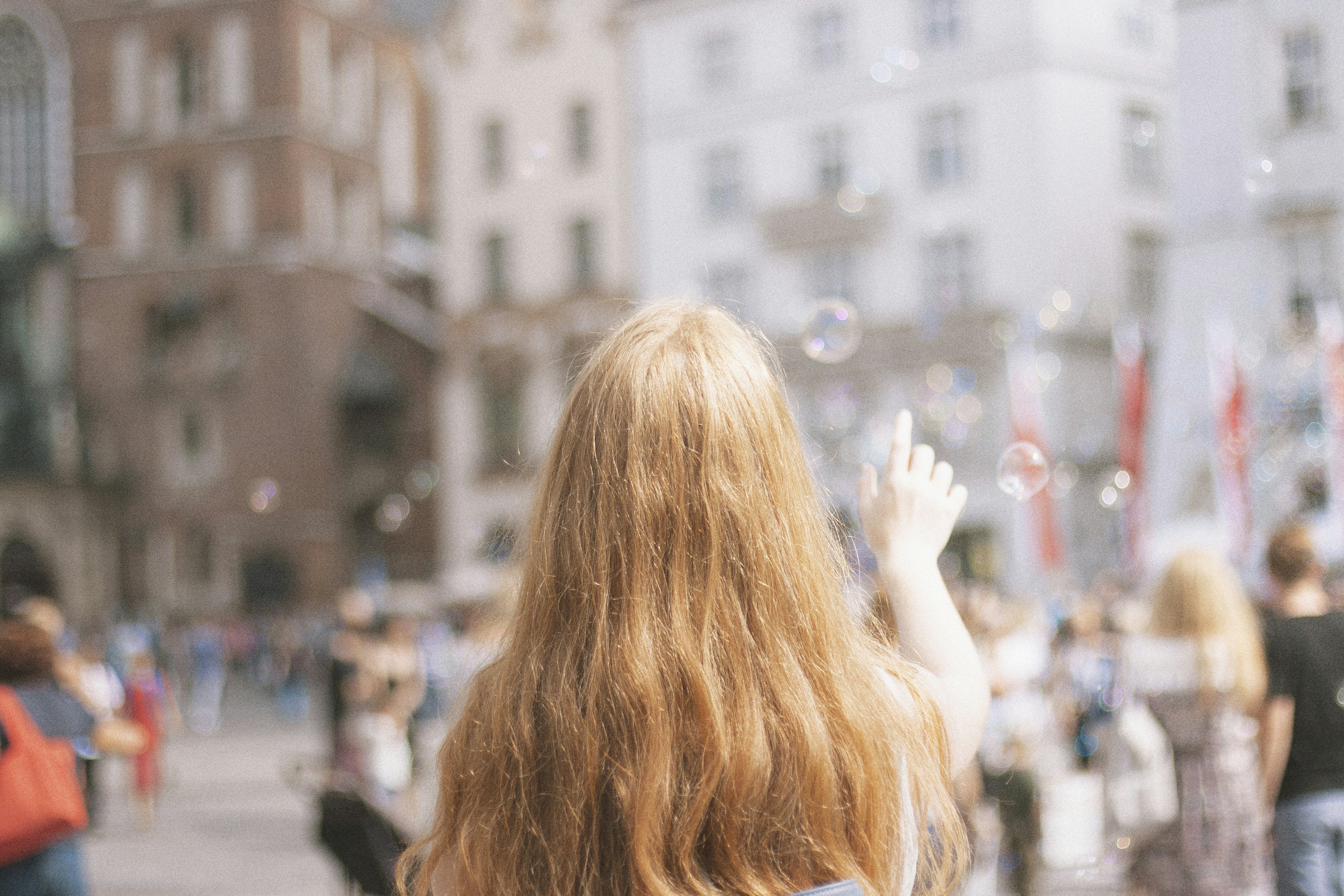 Person with long blonde hair reaching for bubbles in a sunlit city square.