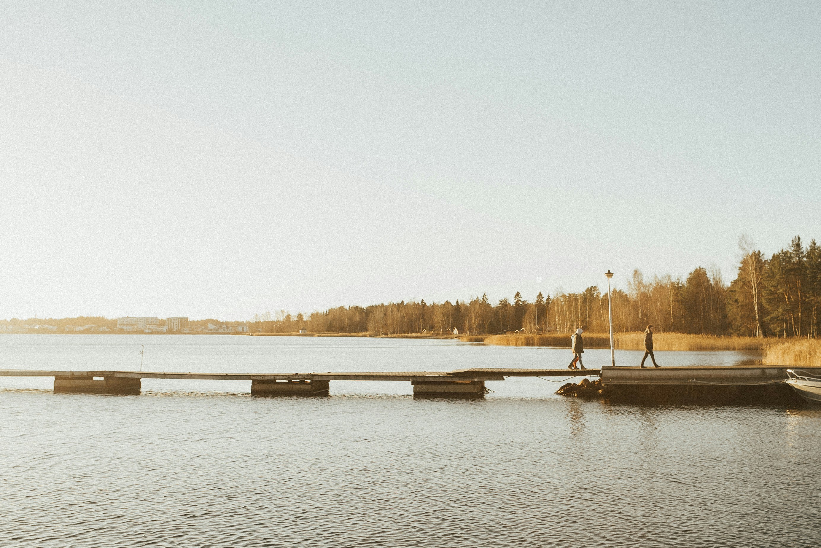 Person walking along a narrow pier stretching across a calm lake under a clear sky.