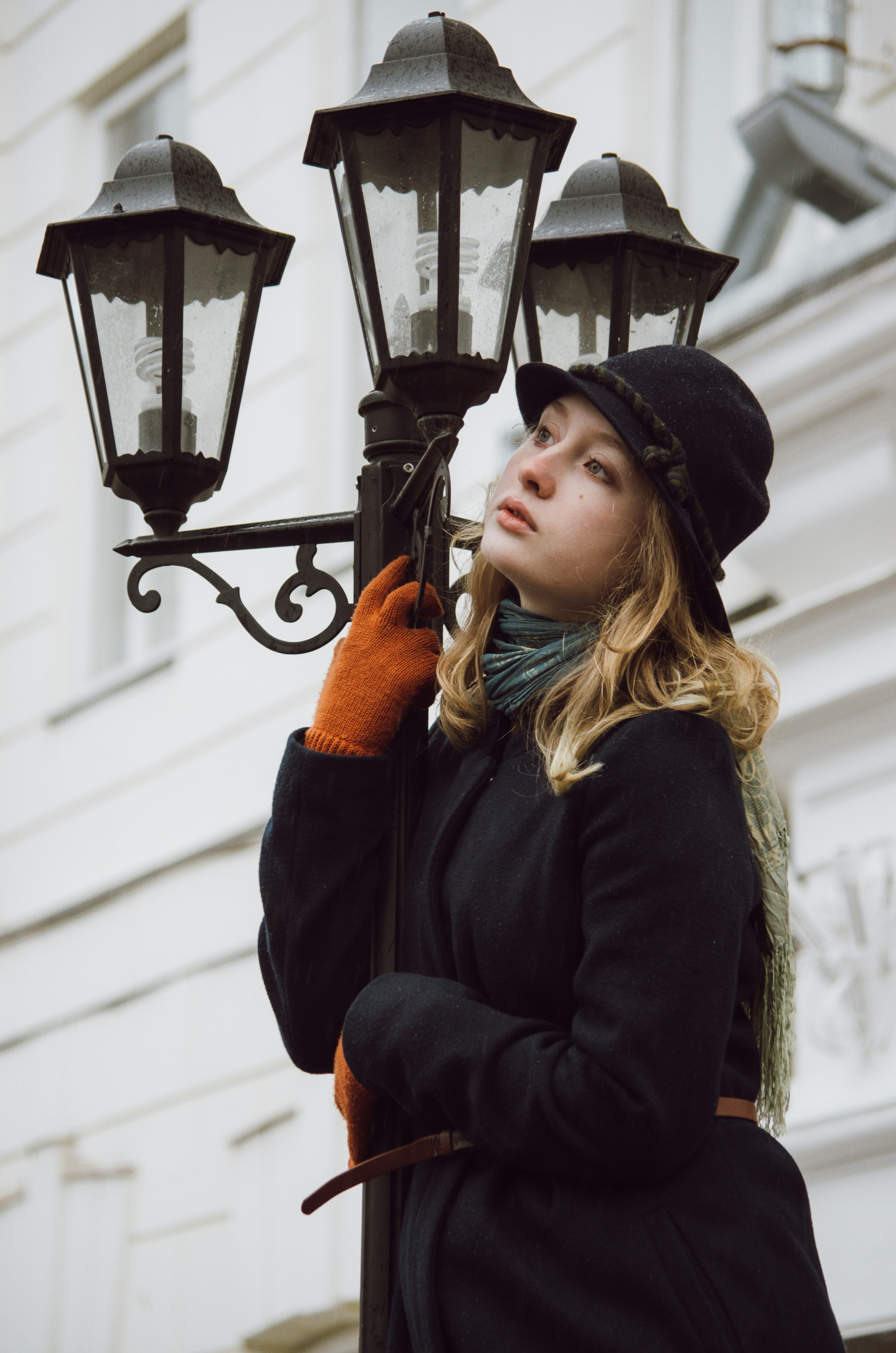 Candid photograph of a woman in a dark coat and hat leaning toward a trio of decorative streetlamps against a pale building.