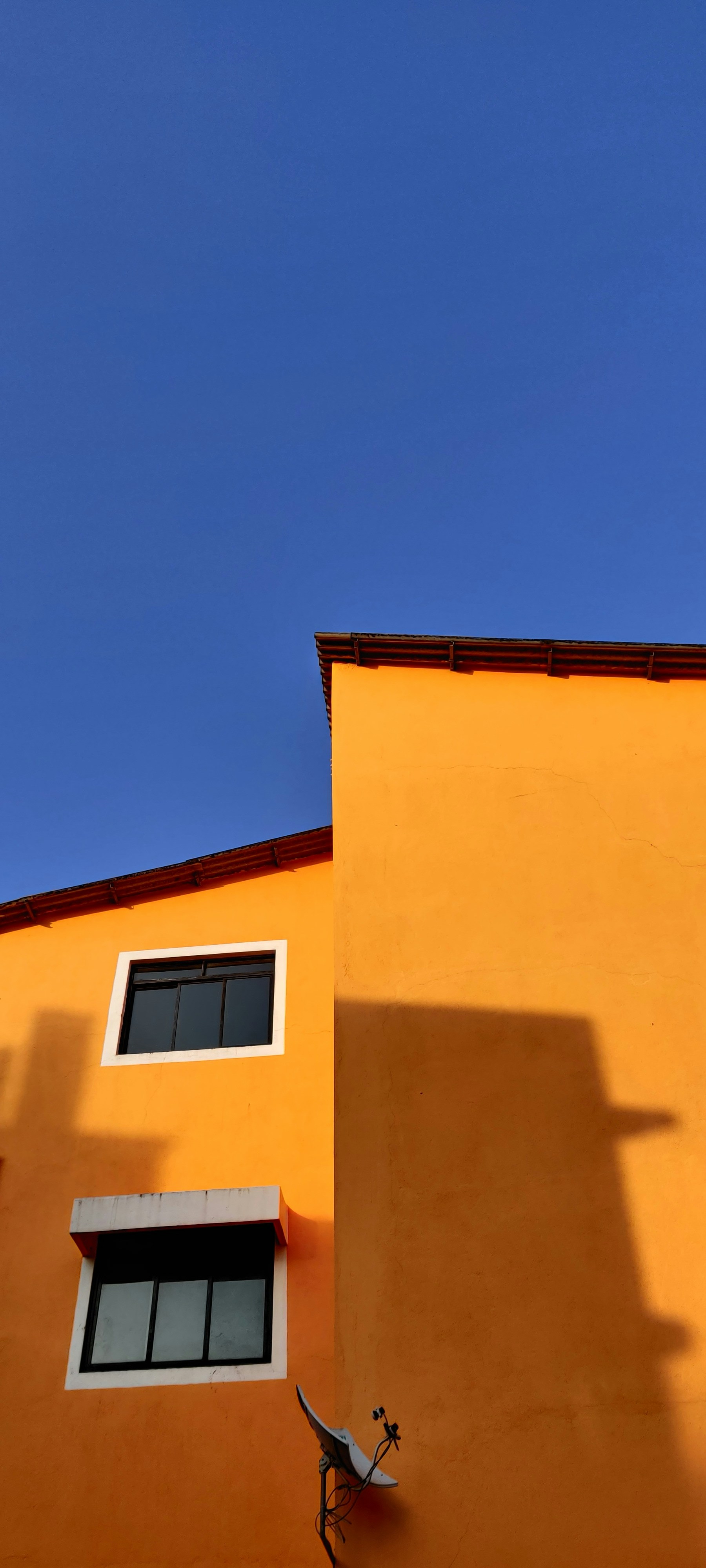 Vibrant orange wall of a building contrasted against a clear blue sky, showcasing geometric shapes and shadows.