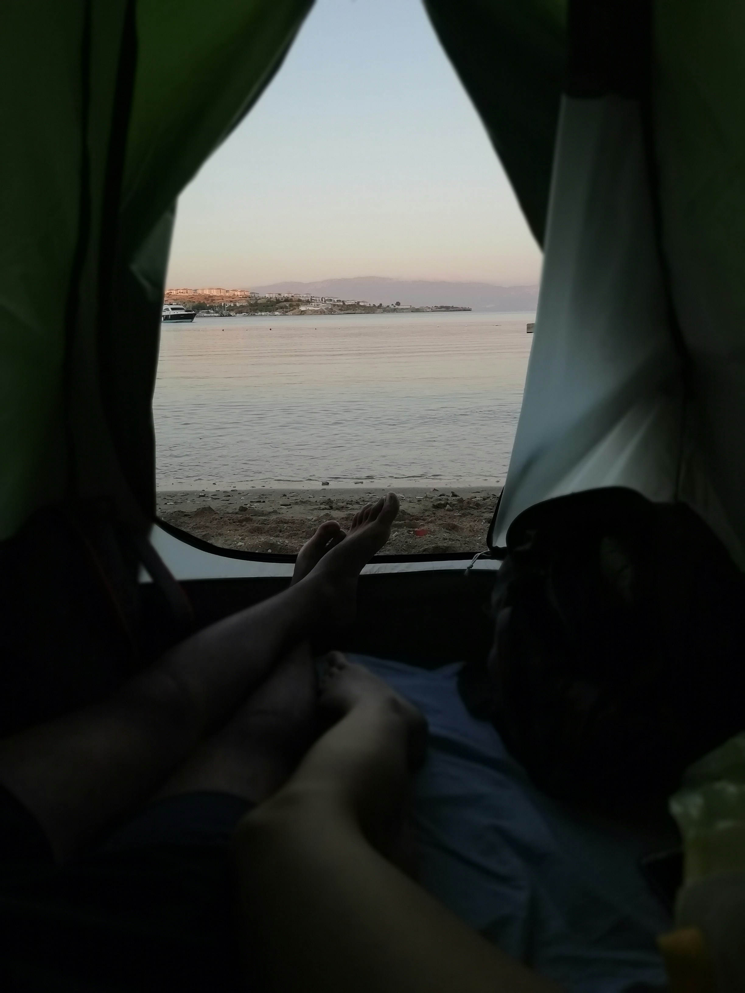 View of a tranquil beach at dusk through the opening of a tent, with relaxed feet visible in the foreground. 