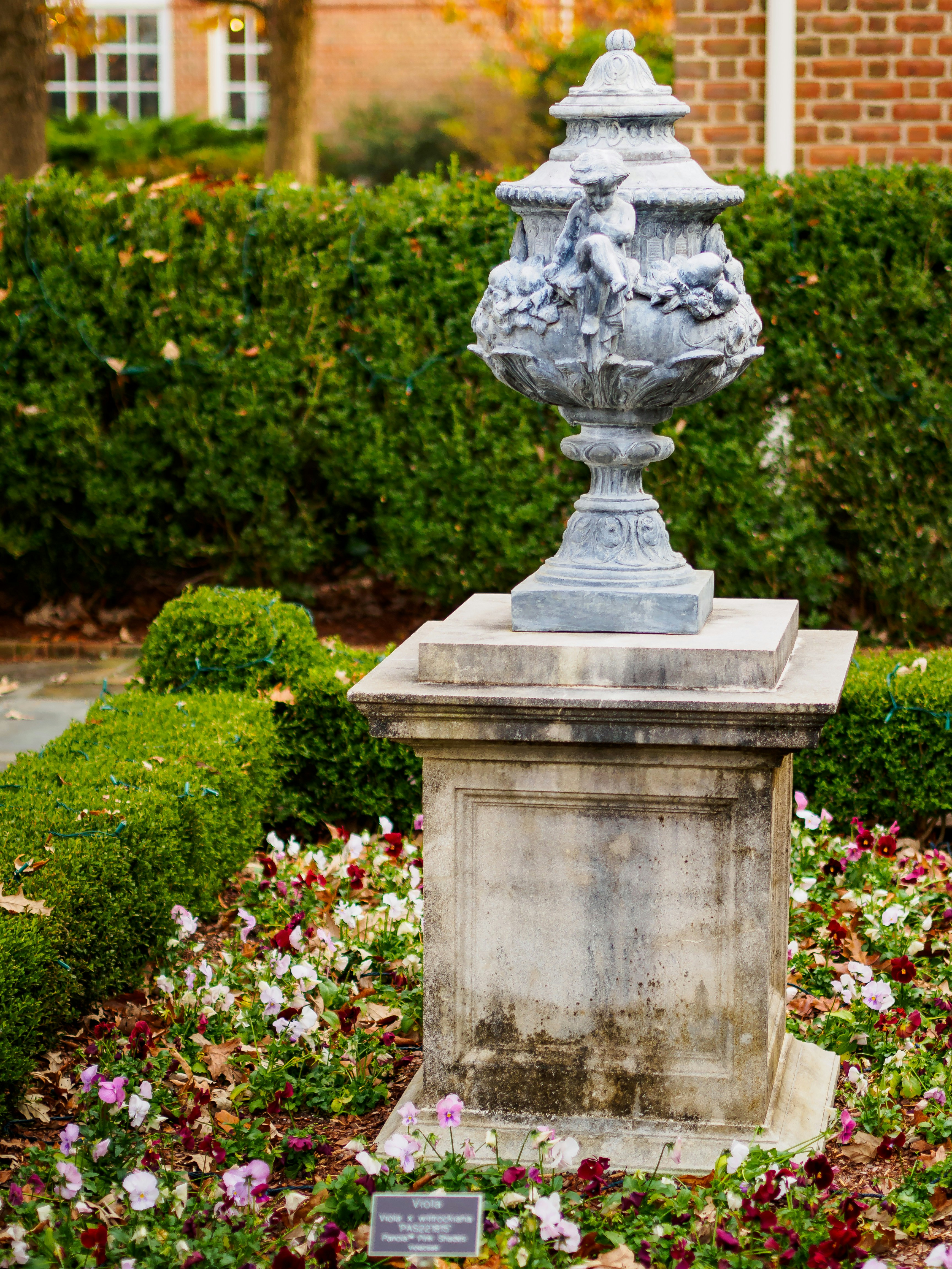 Ornate stone urn atop a pedestal surrounded by vibrant flowers and neatly trimmed hedges.