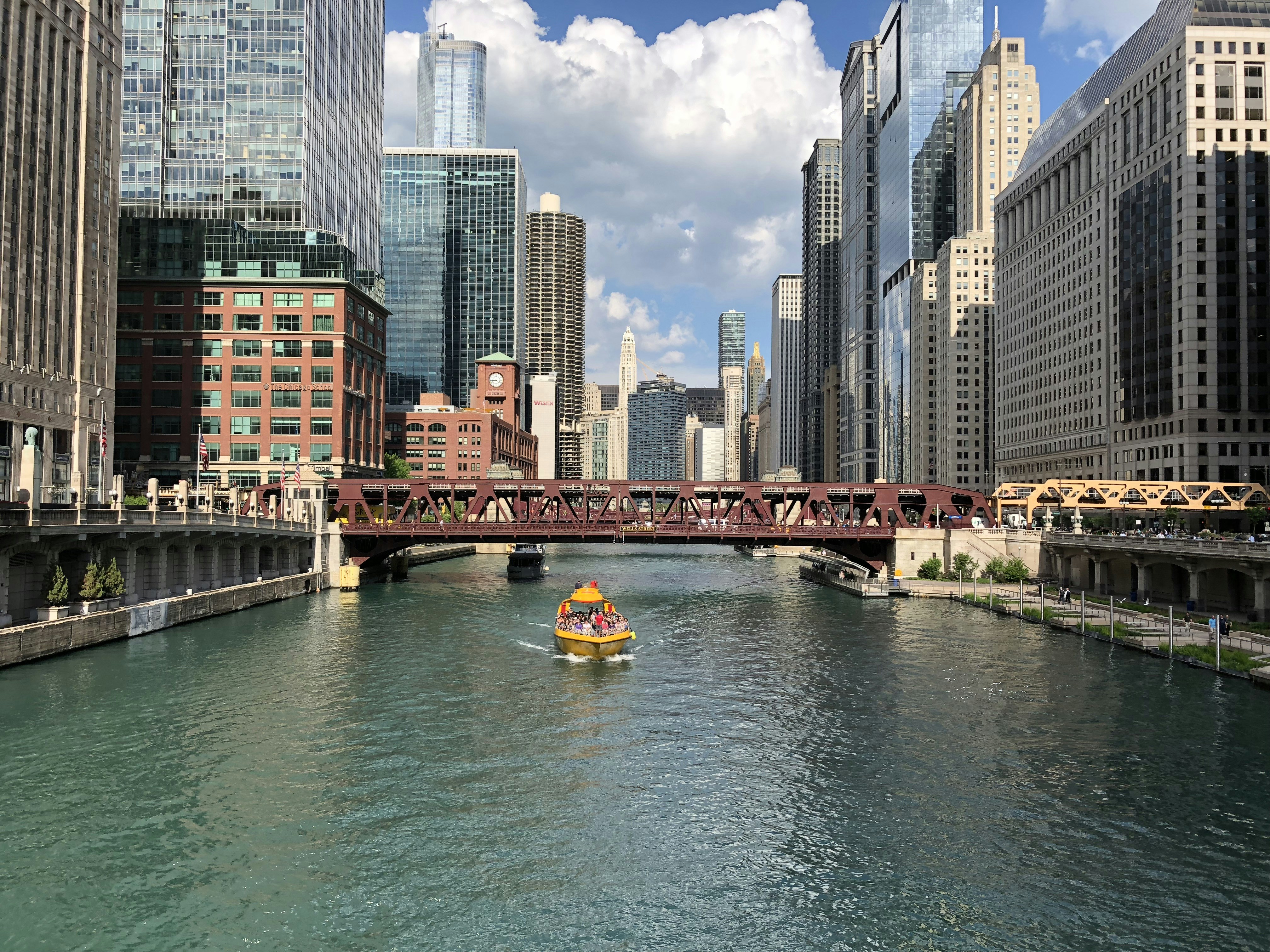 Colorful boat gliding through the Chicago River, flanked by towering skyscrapers and a historic bridge. A vibrant urban scene reflecting the city's architectural diversity.