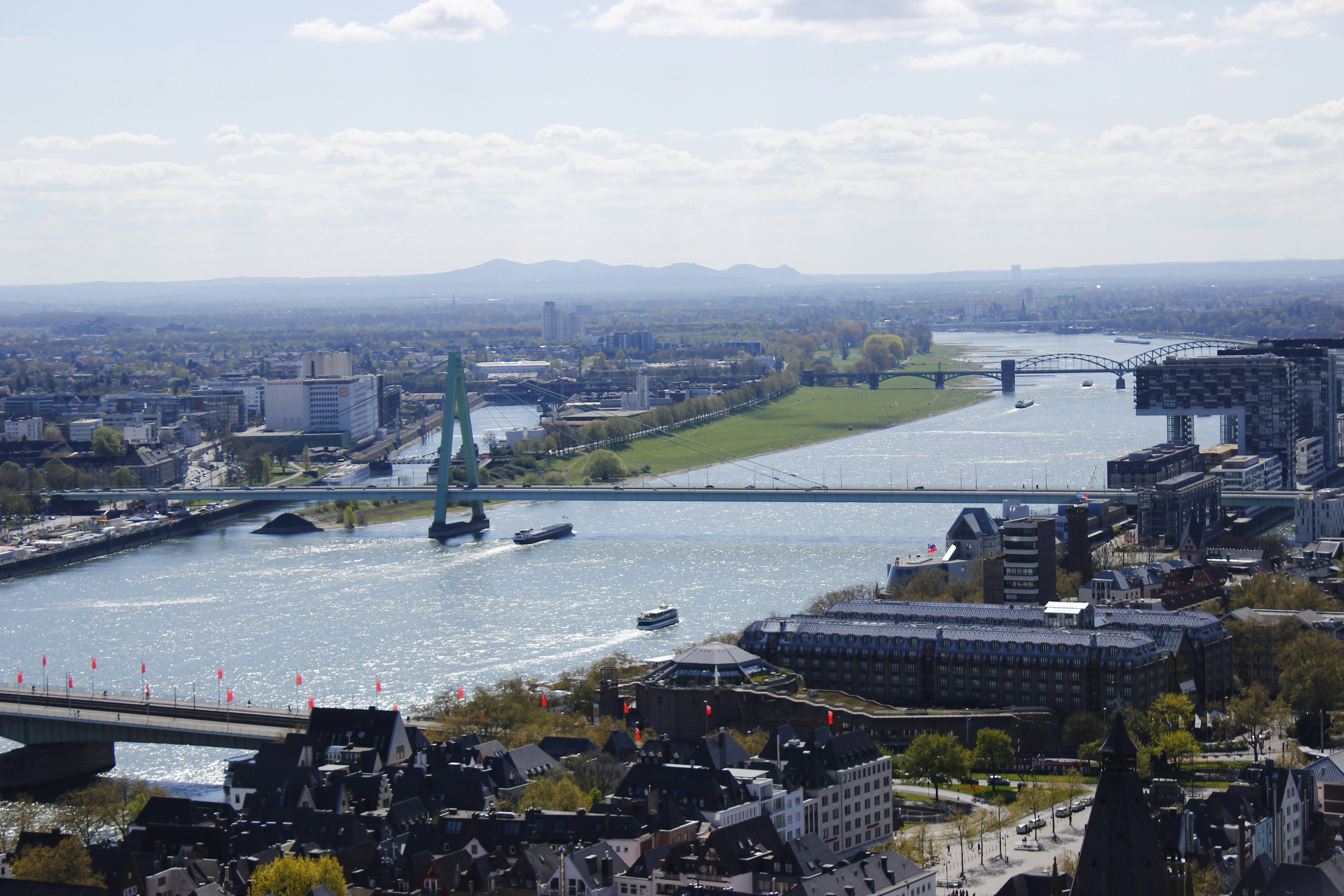 Aerial view of a gray concrete bridge spanning a river, surrounded by urban architecture and green spaces under a partly cloudy sky.