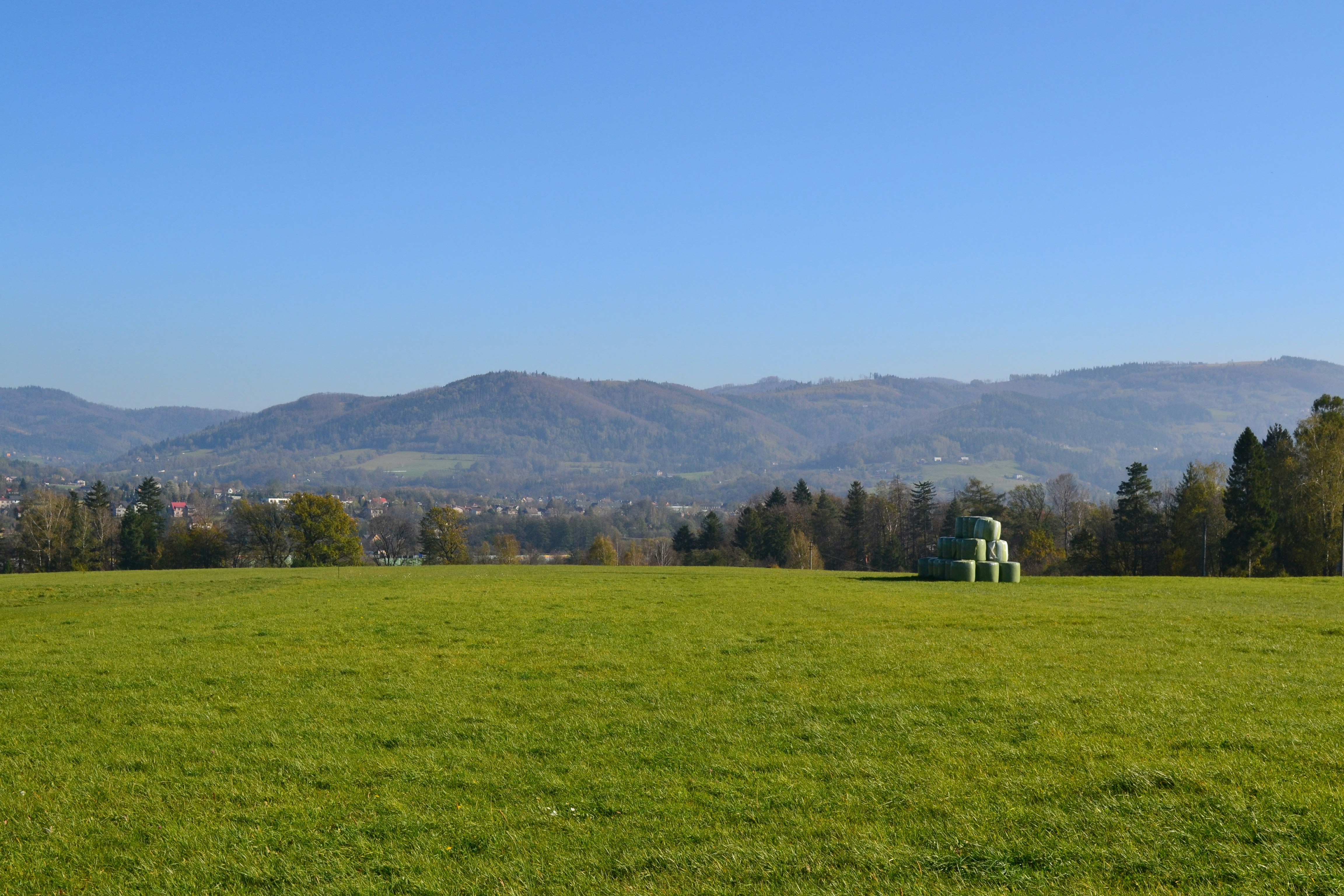 Rolling green fields stretch towards distant mountains under a clear blue sky, with hay bales neatly stacked in the foreground.