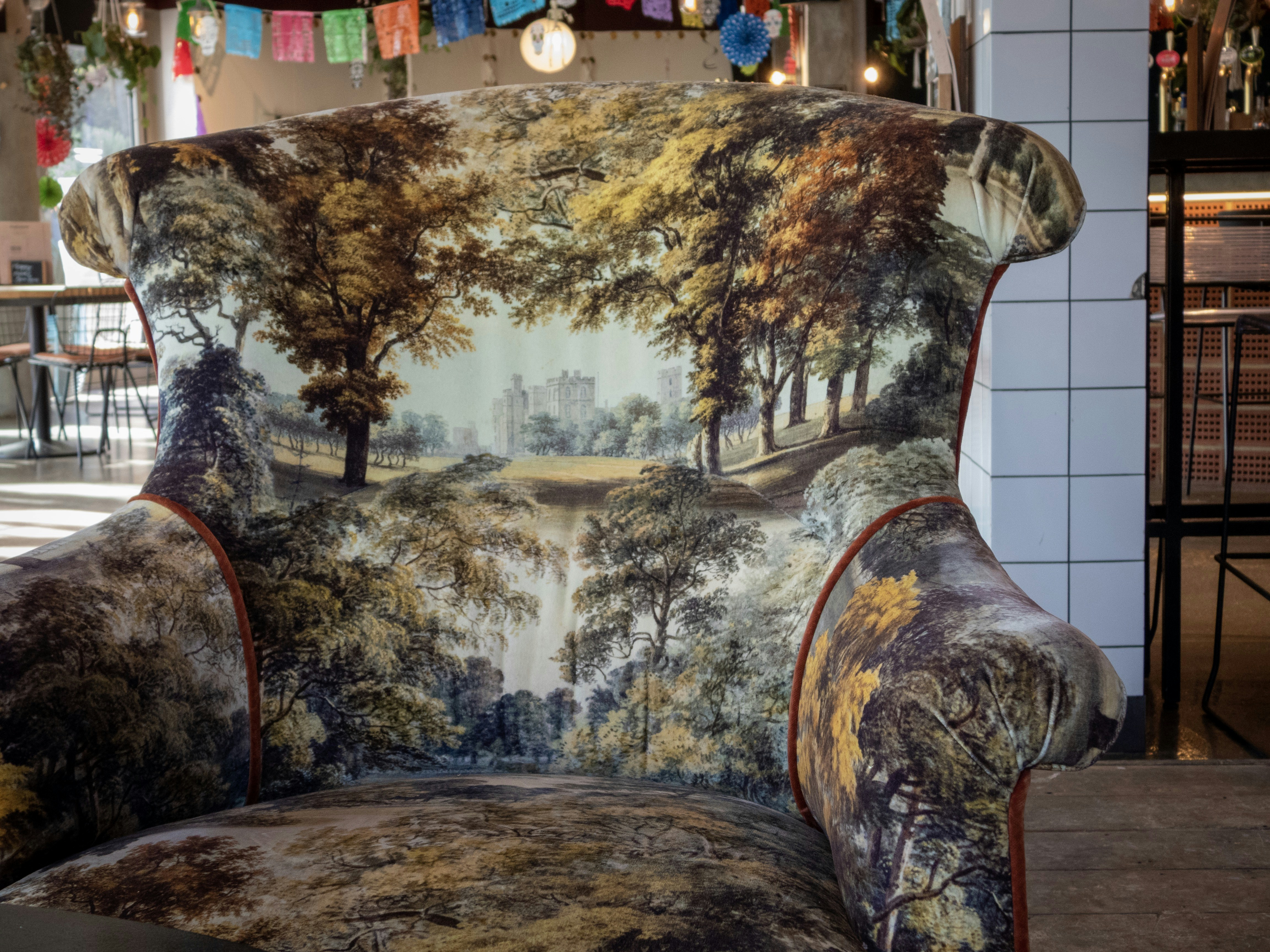 A vintage armchair adorned with a scenic landscape fabric, set in a lively café environment. The intricate design invites viewers to relax and explore its charm.