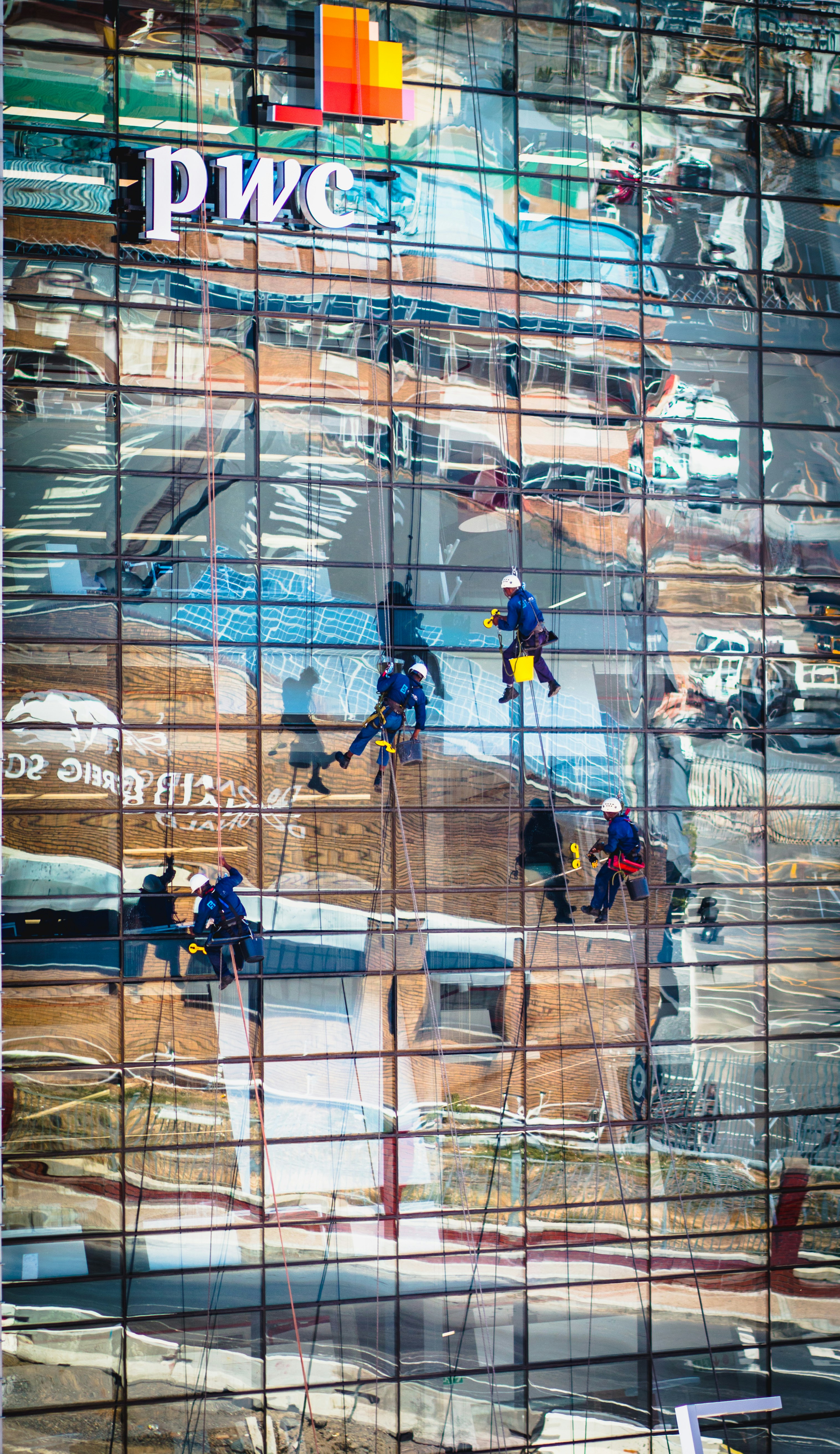 Window cleaners suspended on ropes, meticulously working on a glass facade, while reflections of the cityscape create a layered visual narrative.