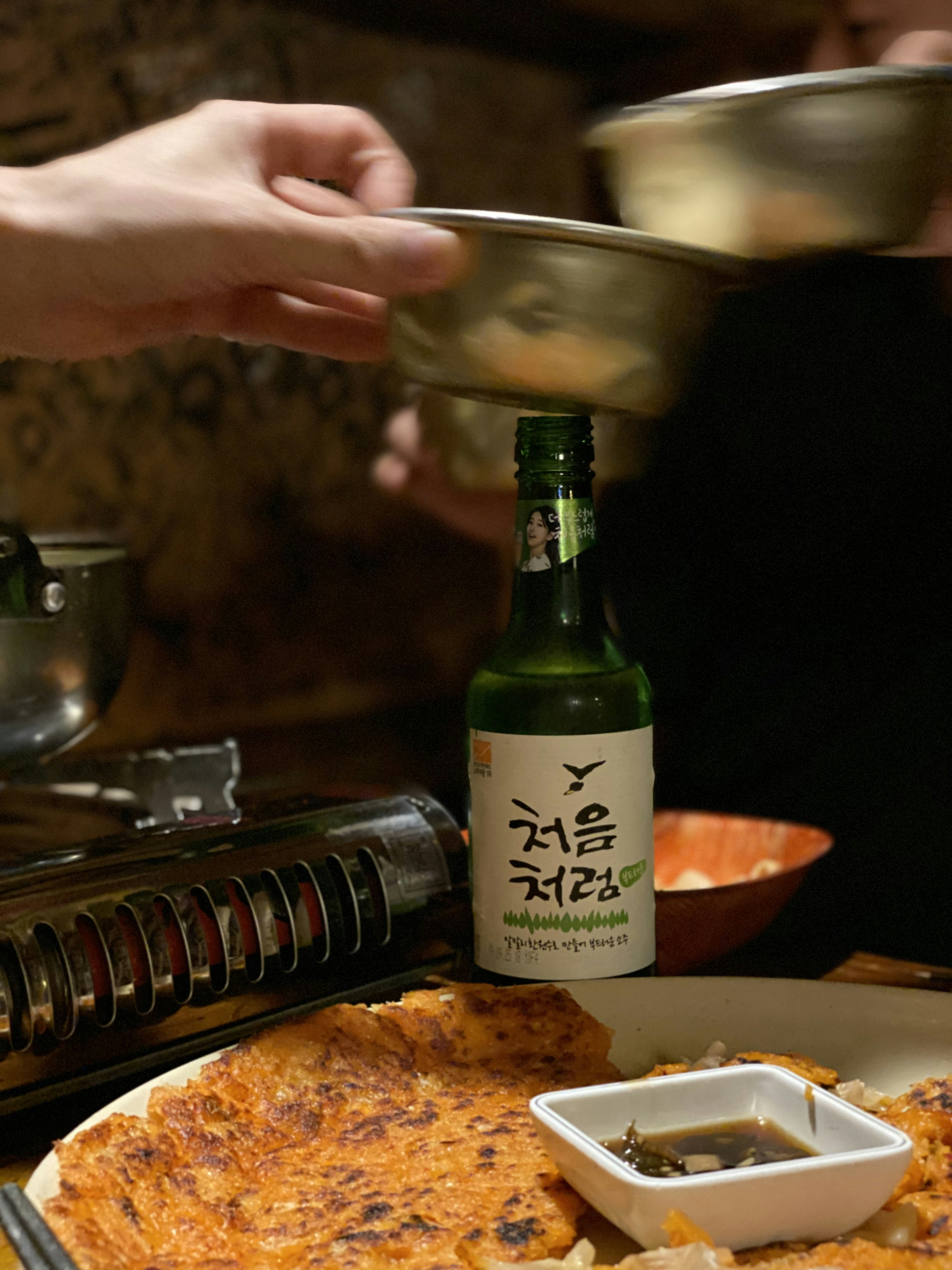 A hand serves a bowl while a bottle of Korean soju stands prominently beside a plate of savory pancake, highlighting a vibrant dining experience.