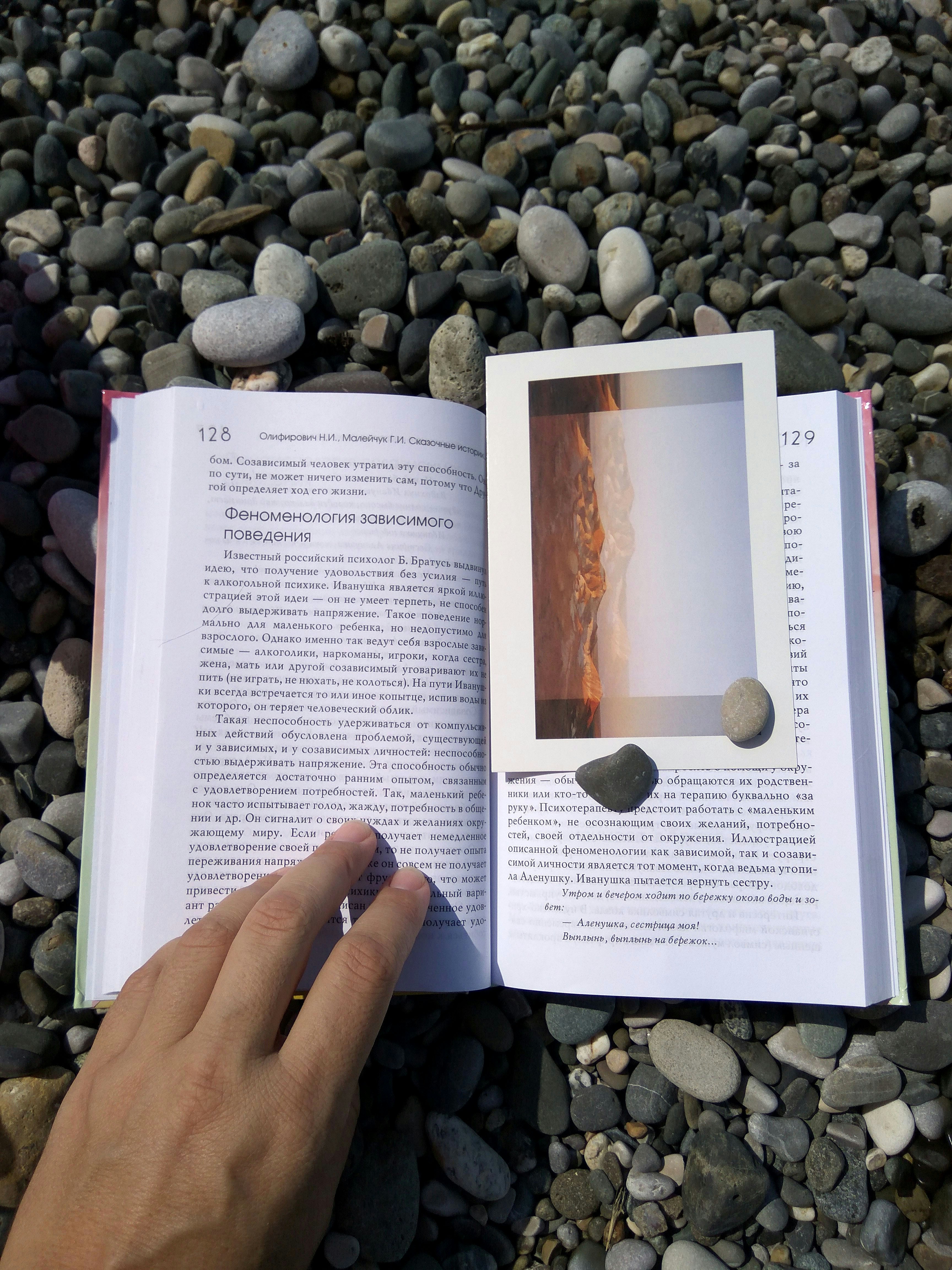 Open book resting on a bed of pebbles, with a photograph placed on top. A hand gently touches the pages, inviting contemplation.