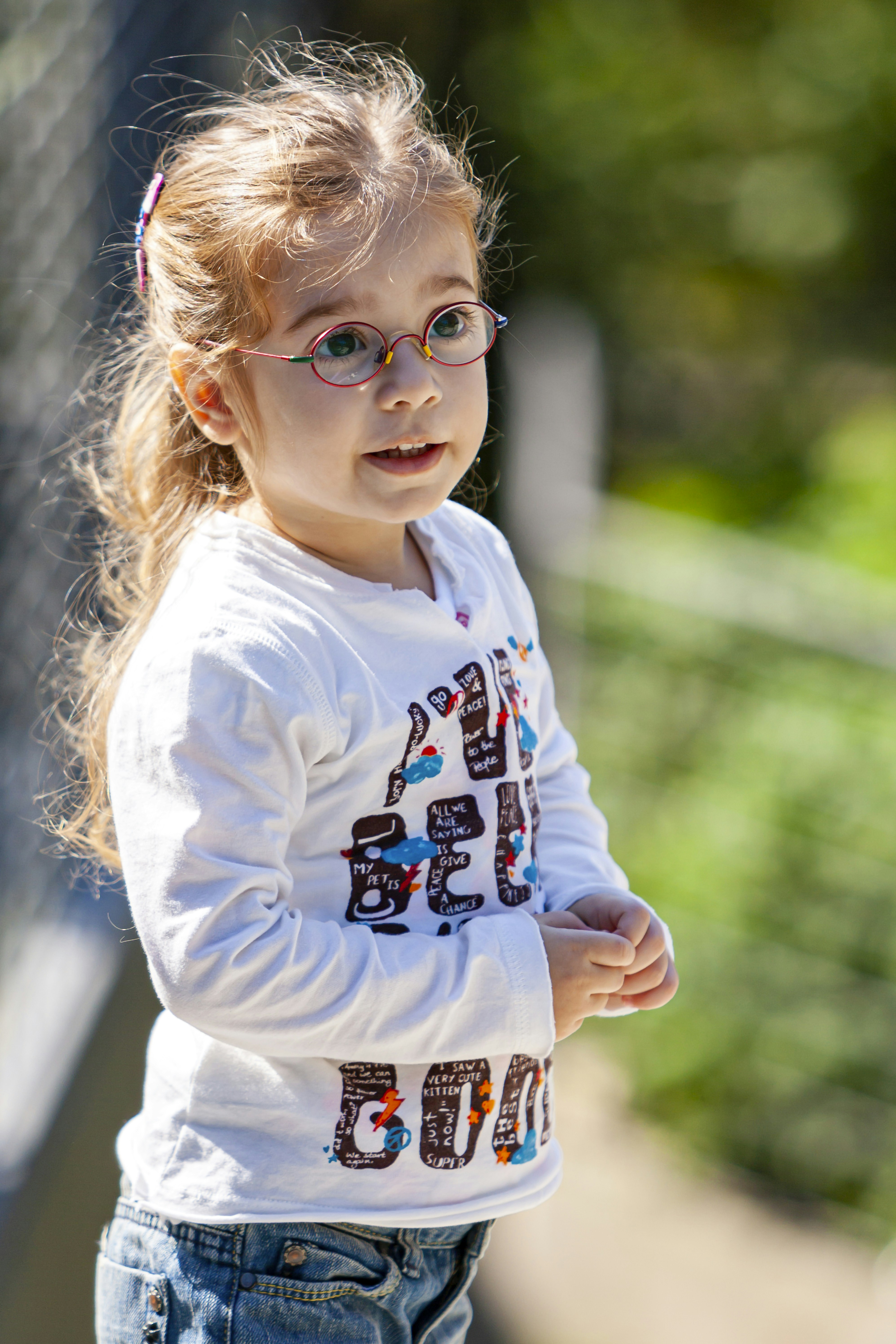 Young girl with glasses smiles while wearing a playful graphic shirt, set against a blurred outdoor background.