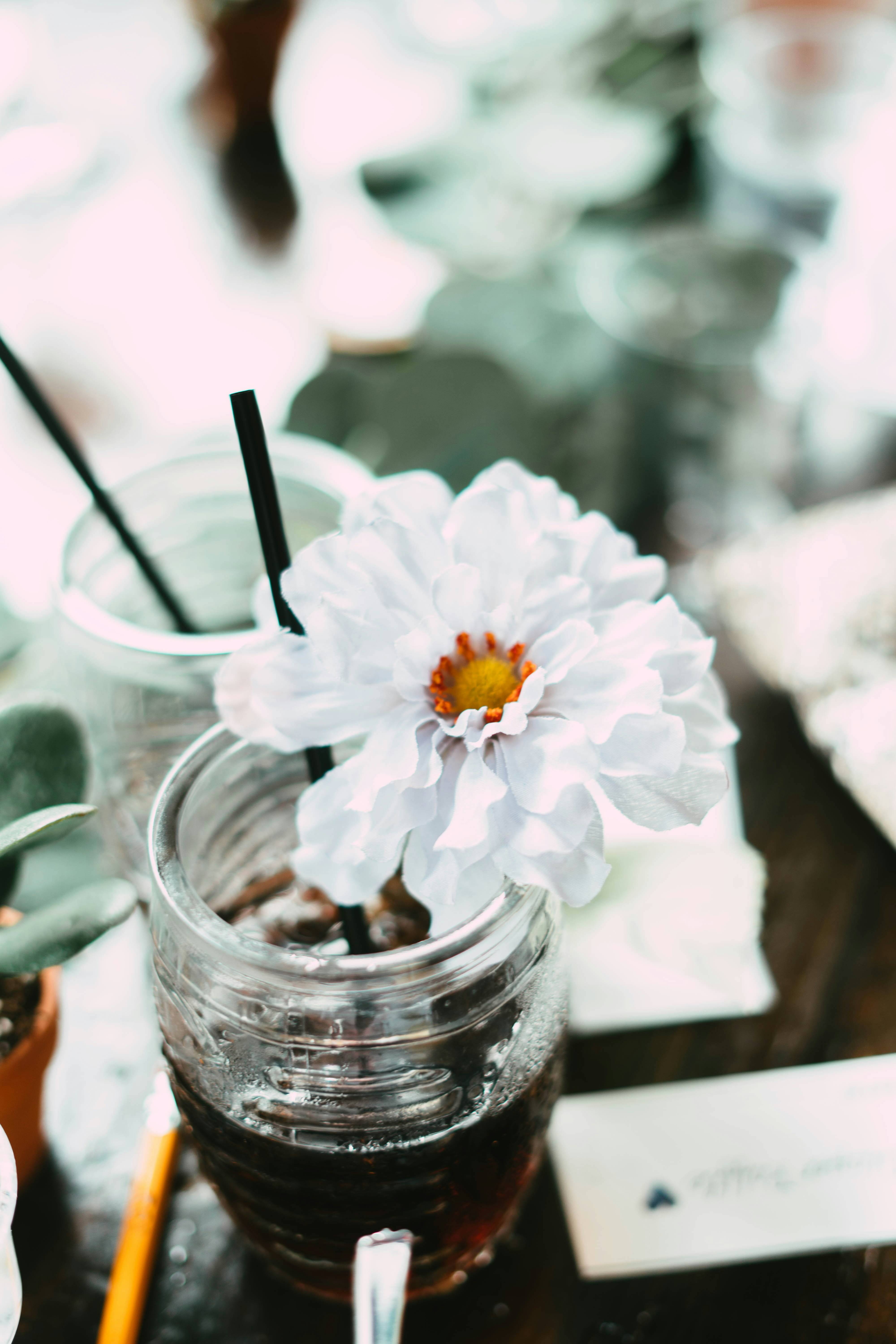 White flower placed in a glass jar with a straw, surrounded by various objects on a wooden table.