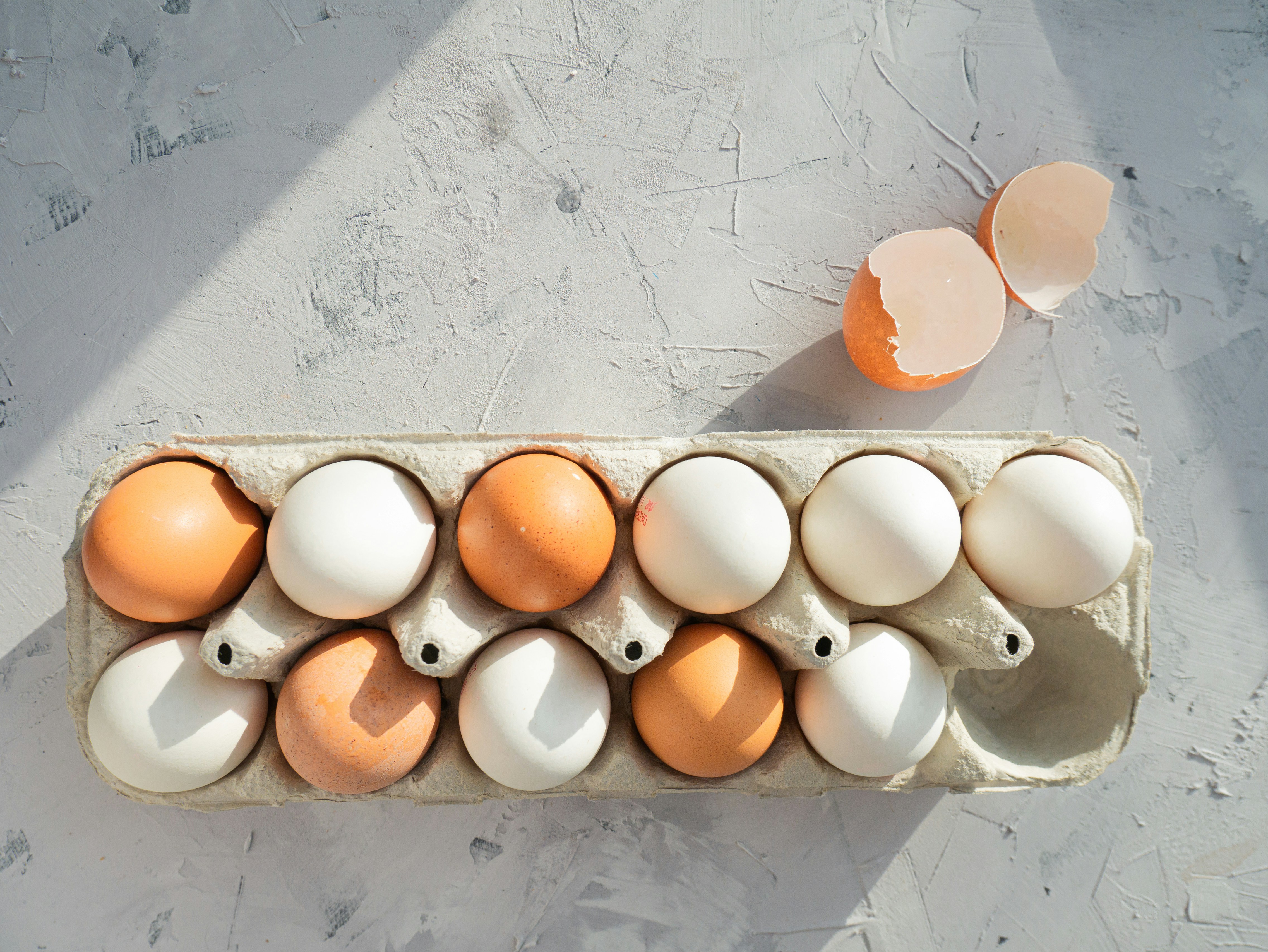 A carton filled with a variety of eggs, showcasing shades of brown and white, with a cracked eggshell lying beside it.