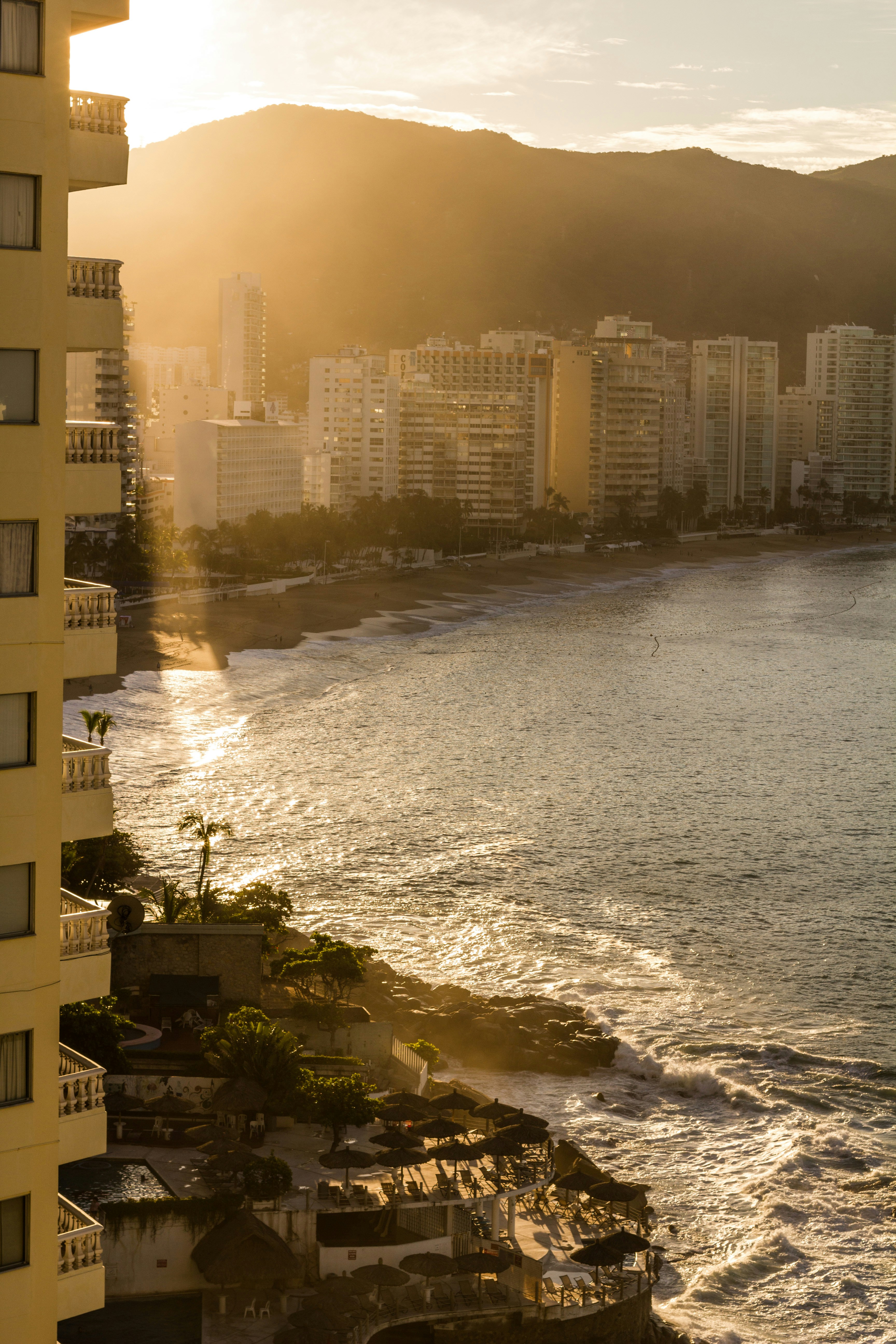 Sunlight glimmers on the ocean waves as urban buildings rise in the background, capturing a tranquil coastal scene at dusk.