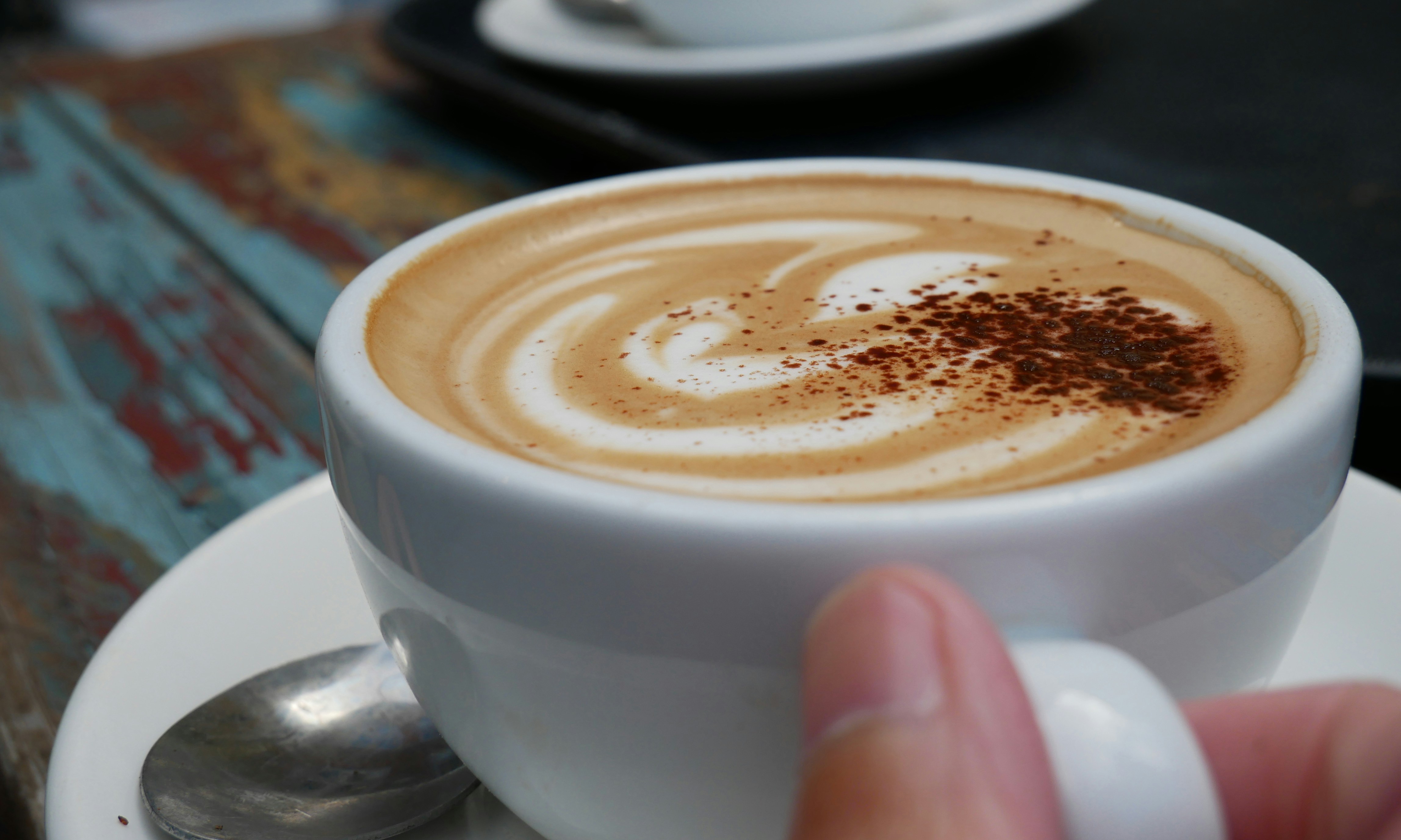 A close-up view of a cappuccino topped with intricate latte art, held by a hand against a rustic table backdrop.