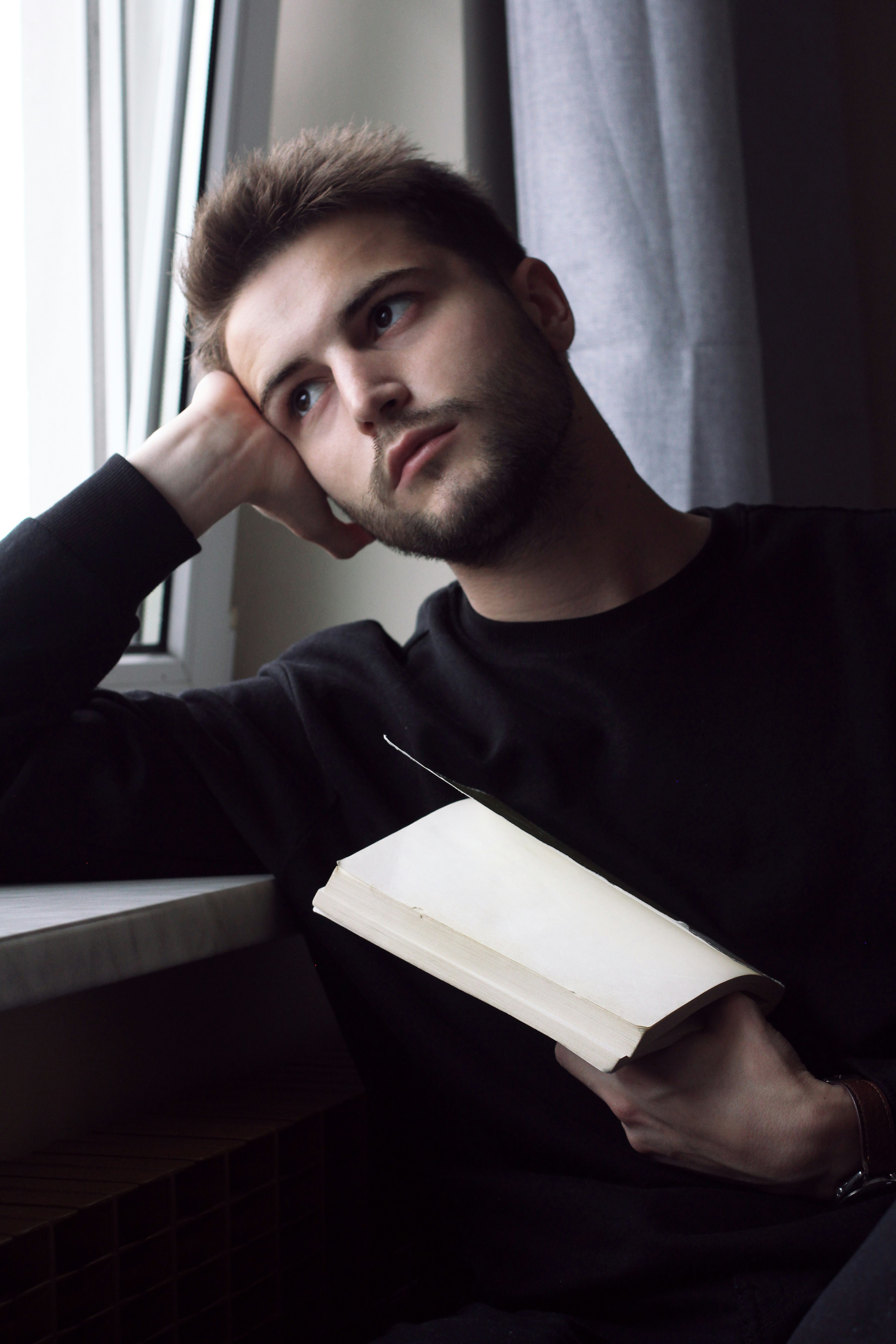 Young man gazing thoughtfully out the window while holding a book, captured in a cozy indoor setting.