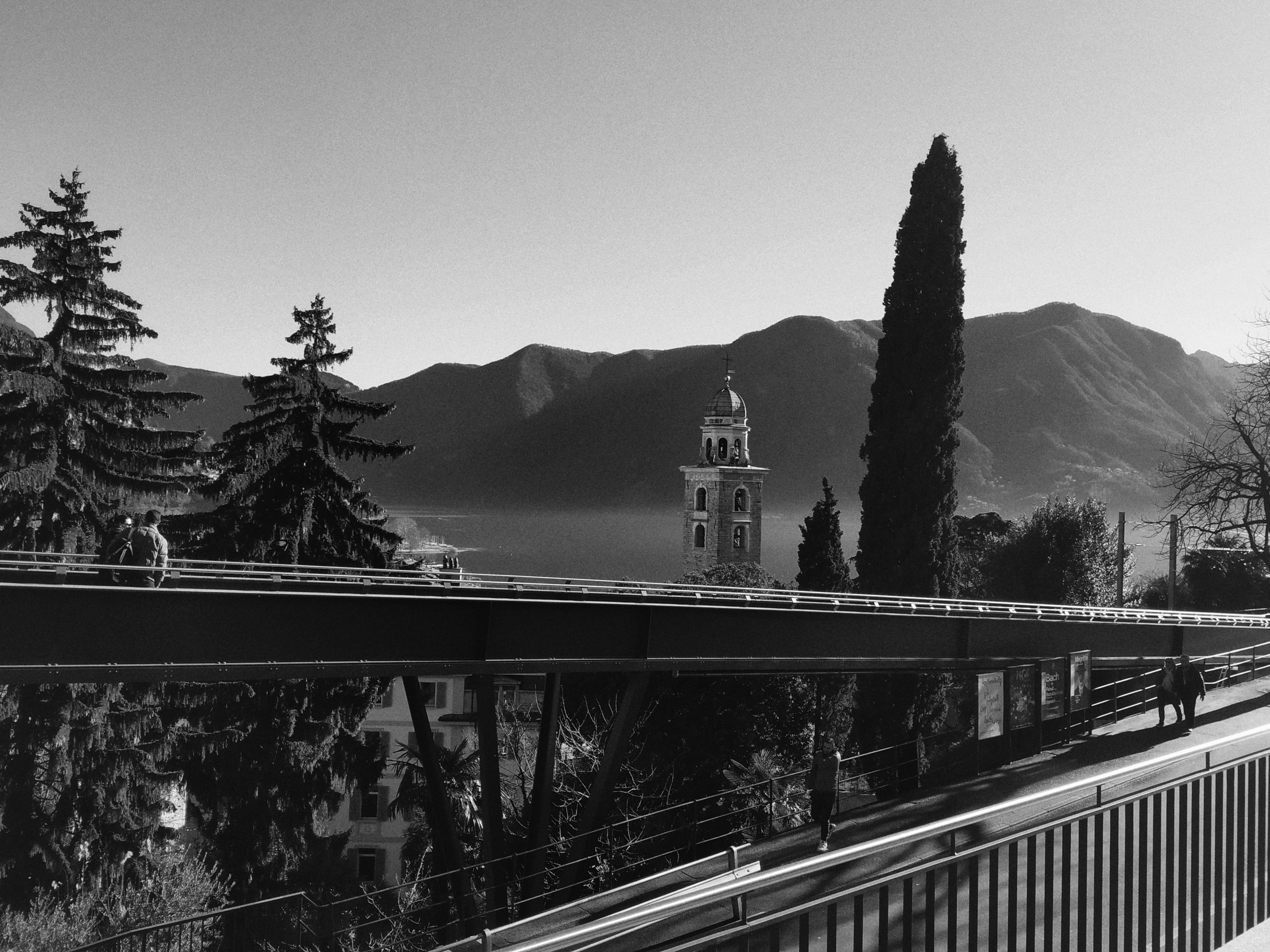 Black-and-white photograph capturing a lakeside town with a tall bell tower, a foreground bridge, and pine trees set against distant mountains.