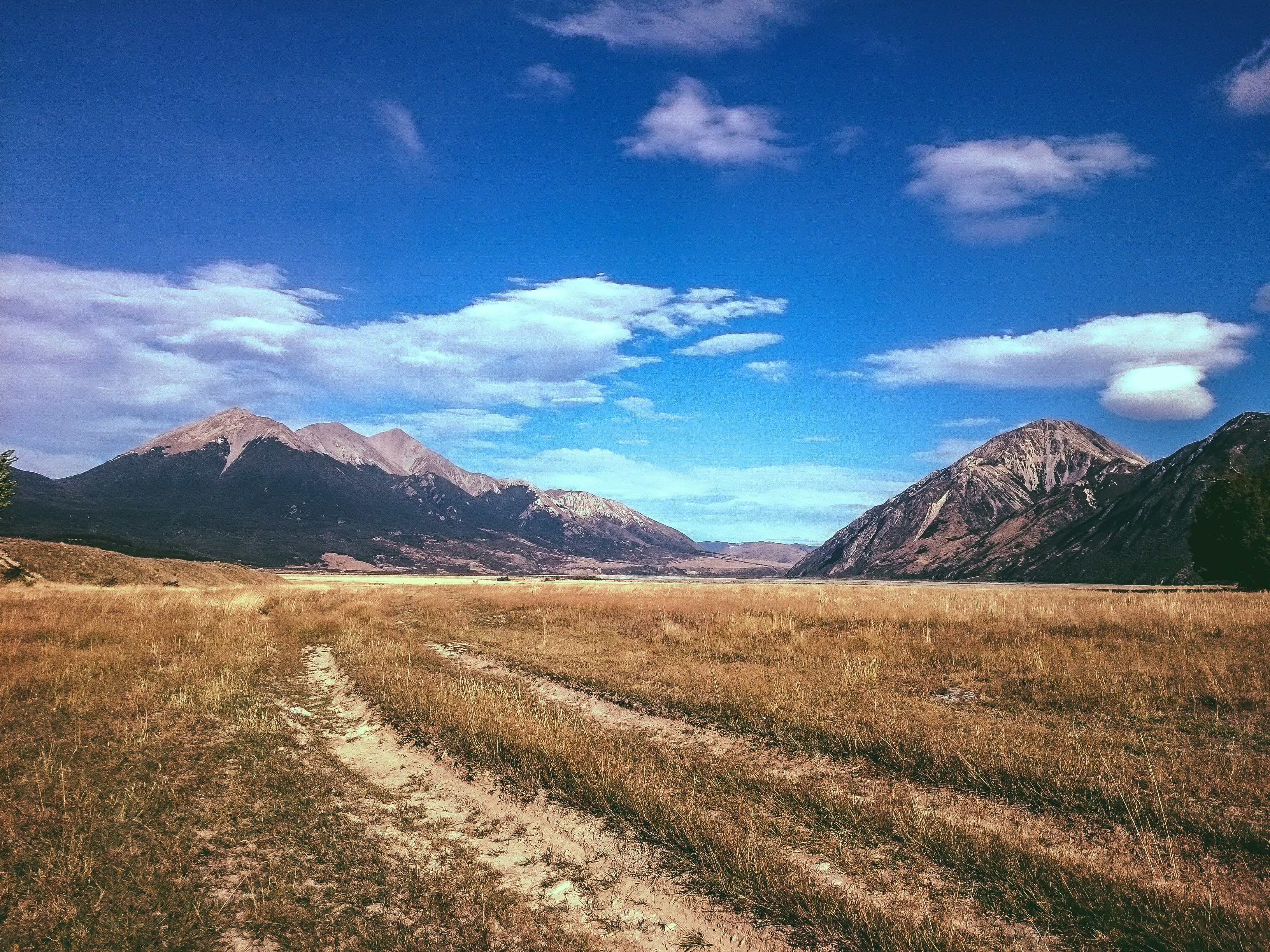 Dirt road meanders through a golden grassy field flanked by rugged mountain ranges under a bright blue sky.