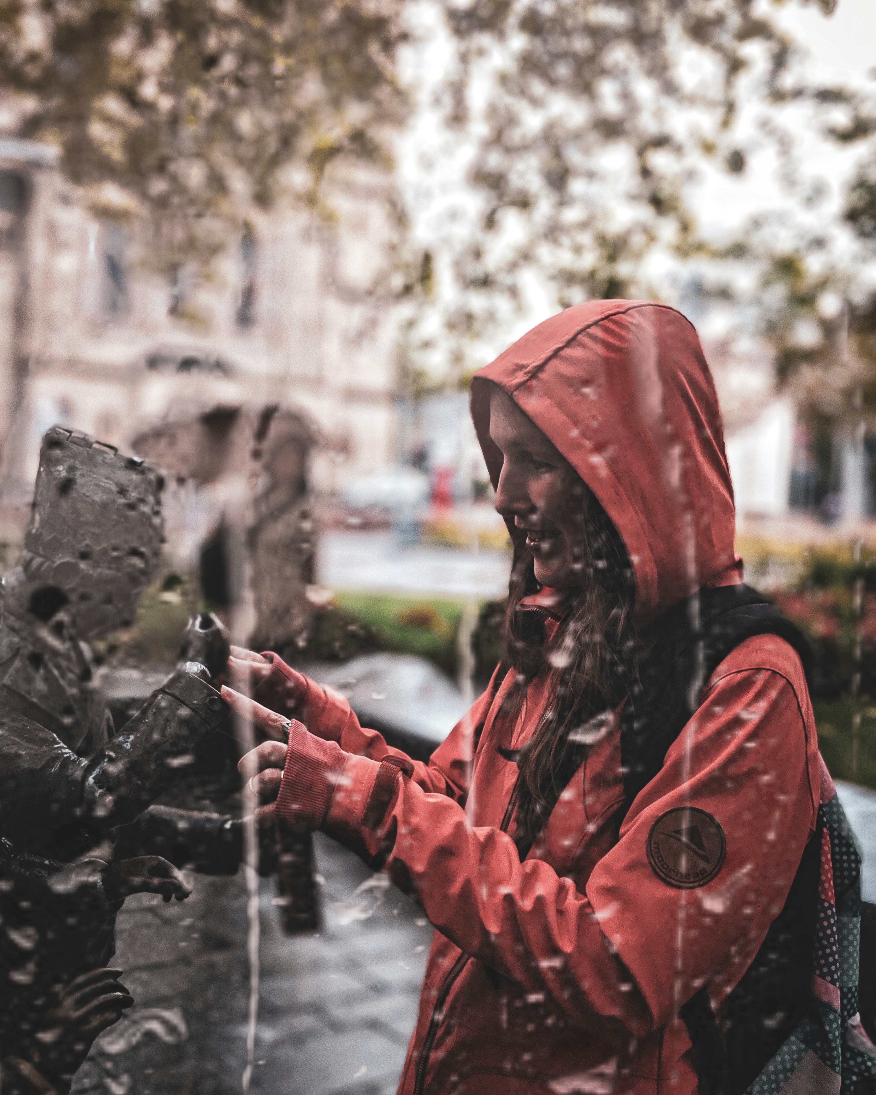 Person in a red hooded jacket interacting with a statue on a rainy day.