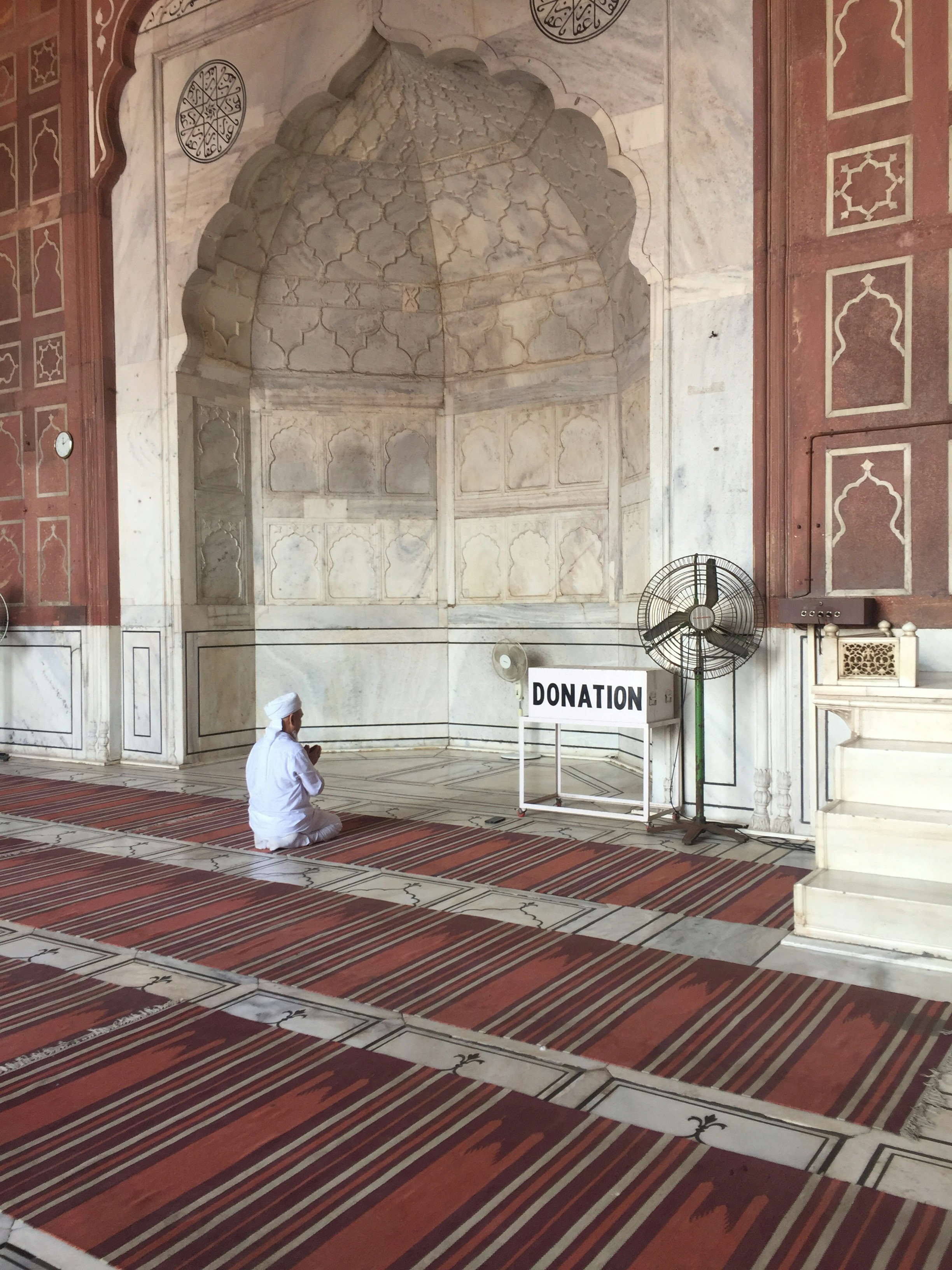 A person in traditional attire kneels in prayer within a beautifully adorned mosque, with a donation box nearby. The intricate architecture and serene atmosphere enhance the spiritual experience.