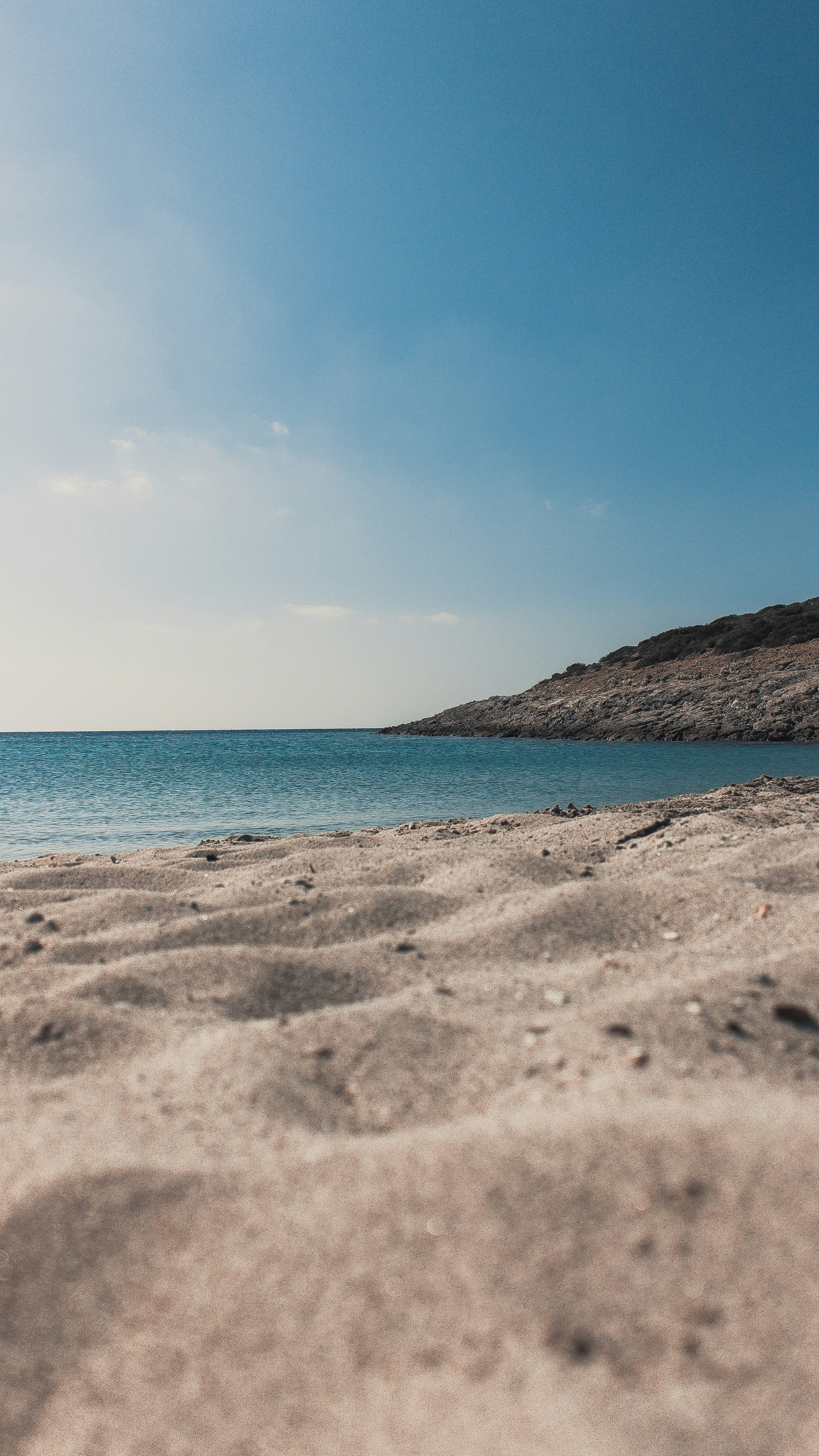Gentle waves caress a sandy beach under a clear blue sky, with a rocky coastline in the distance.