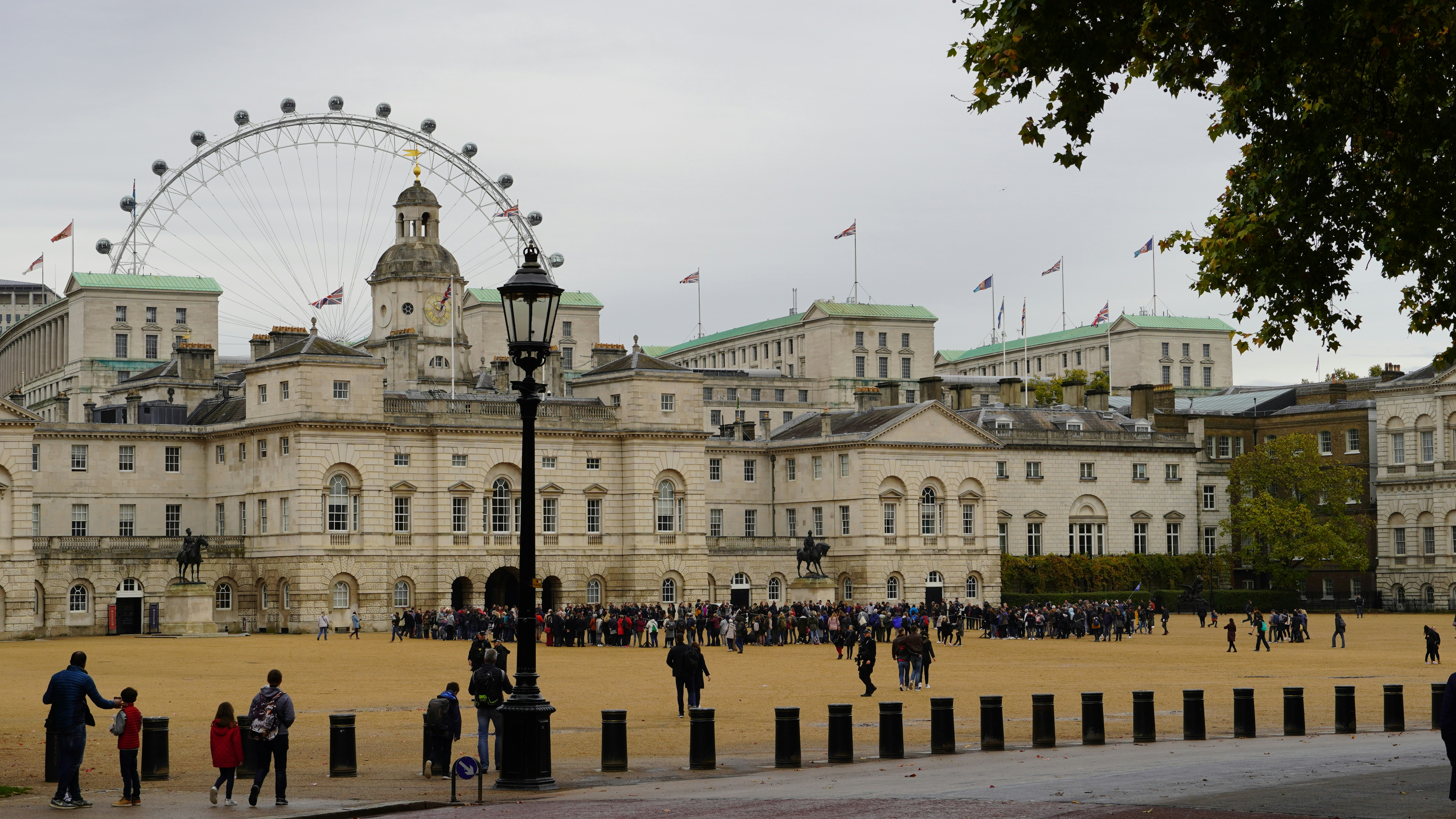 Crowd gathered in front of a historic building with the London Eye in the background, showcasing a blend of architecture and urban life.
