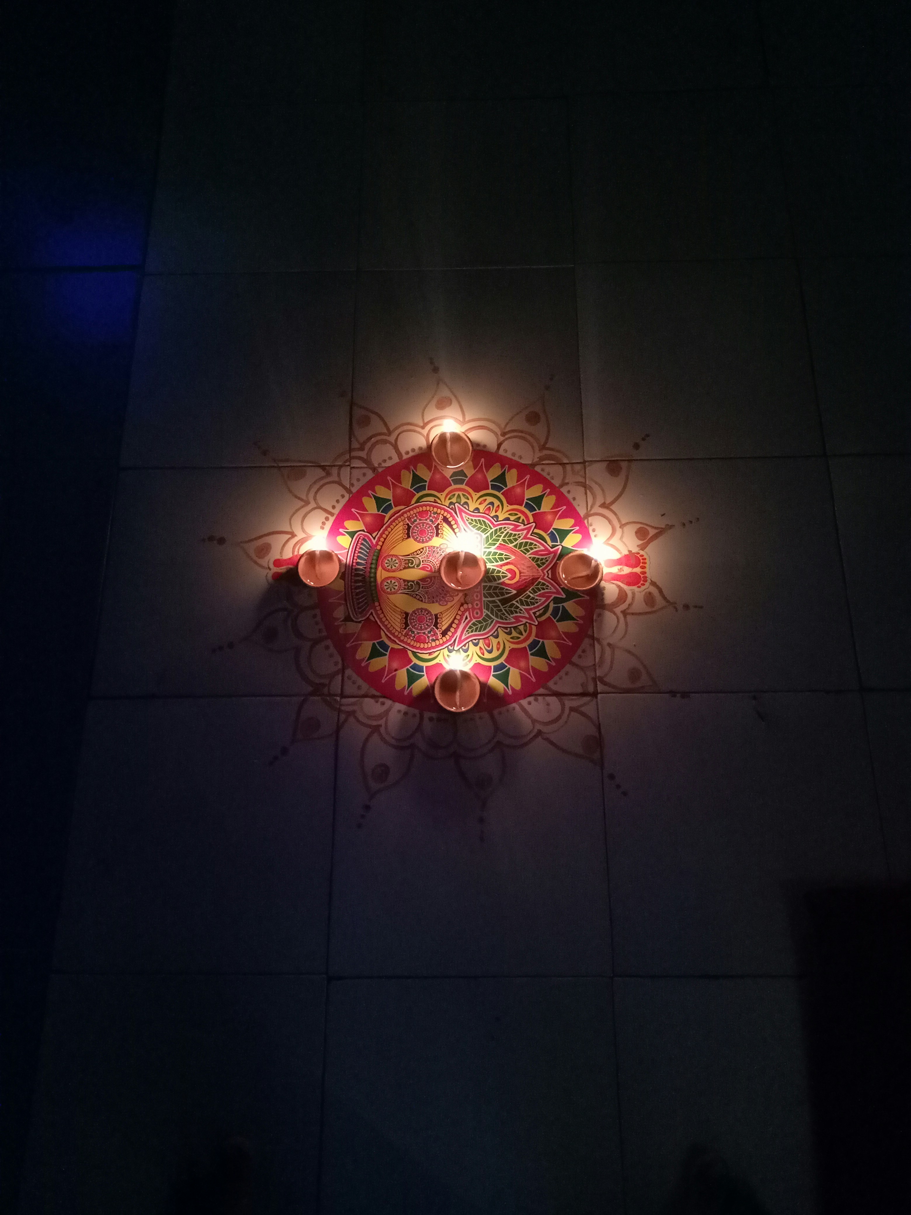 Lit candle mandala glows at the center of a dark tiled floor, its vibrant pattern radiating outward.