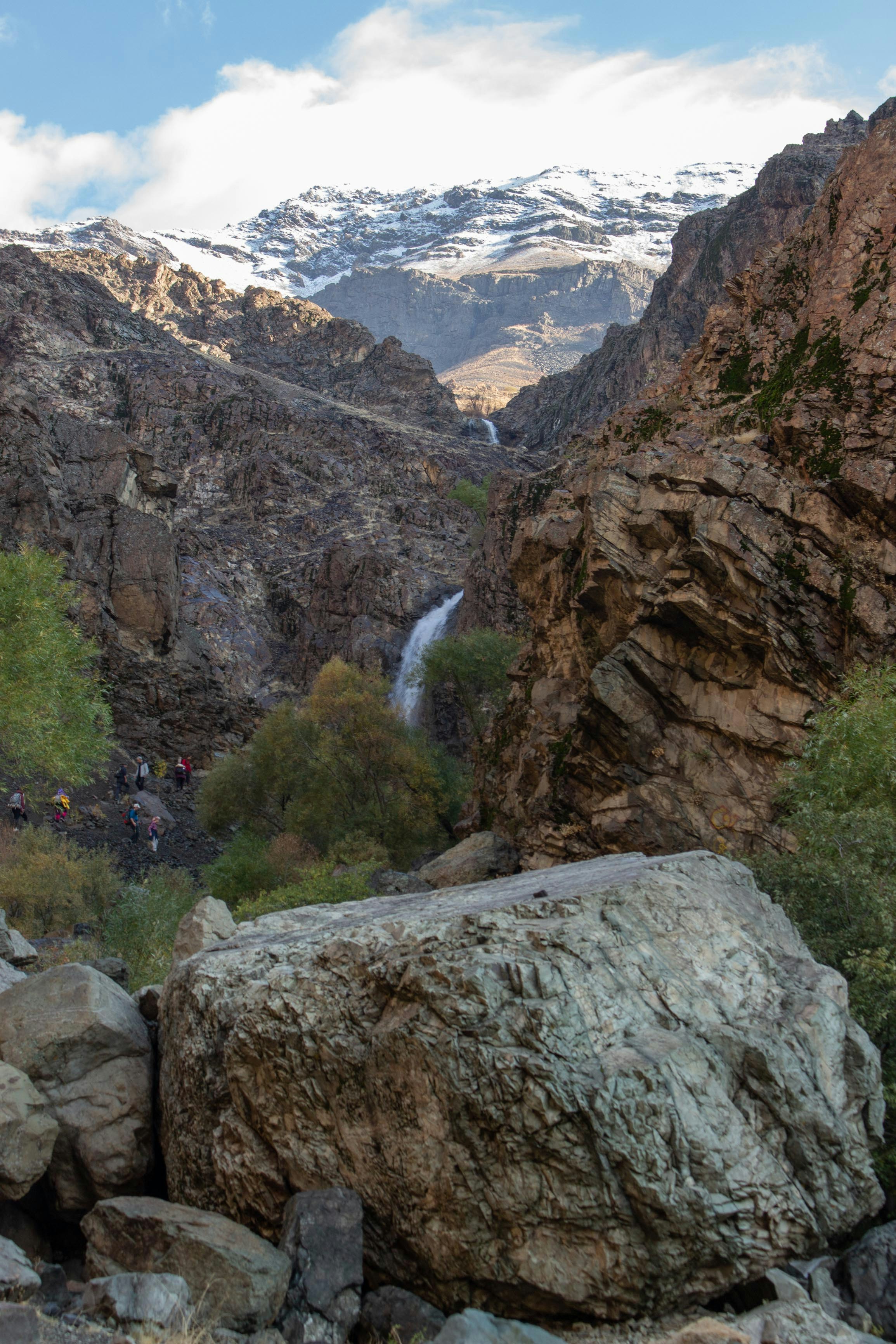 A serene waterfall cascades down rocky cliffs, surrounded by lush greenery and towering mountains. Hikers traverse the rugged terrain, adding a sense of adventure.