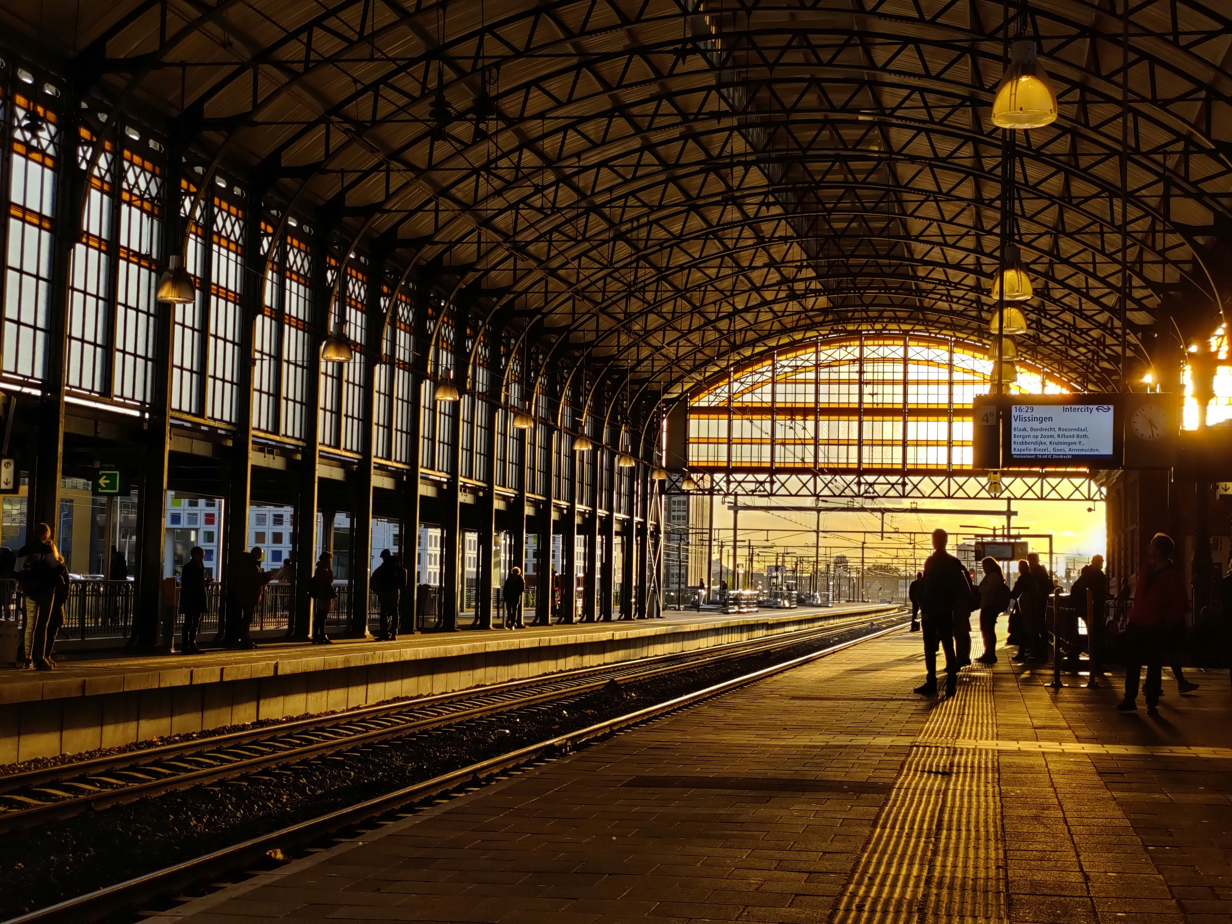 Silhouetted travelers walk along a sunlit train platform beneath an arched ceiling.