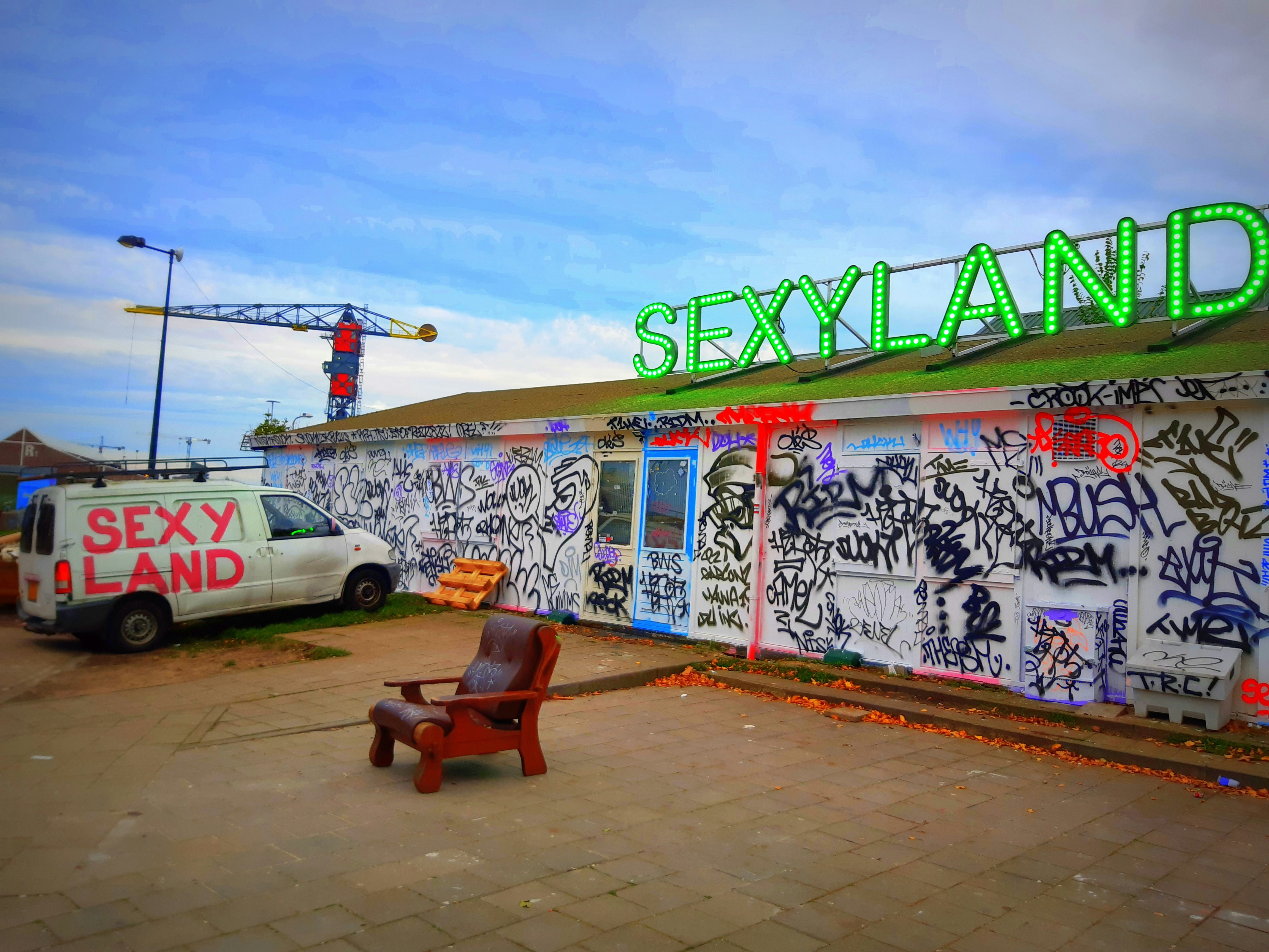 Graffiti-covered storefront photograph dominated by a bright neon 'SEXYLAND' sign. A marked van and a solitary chair sit in the foreground of the expansive, graffiti-strewn lot.