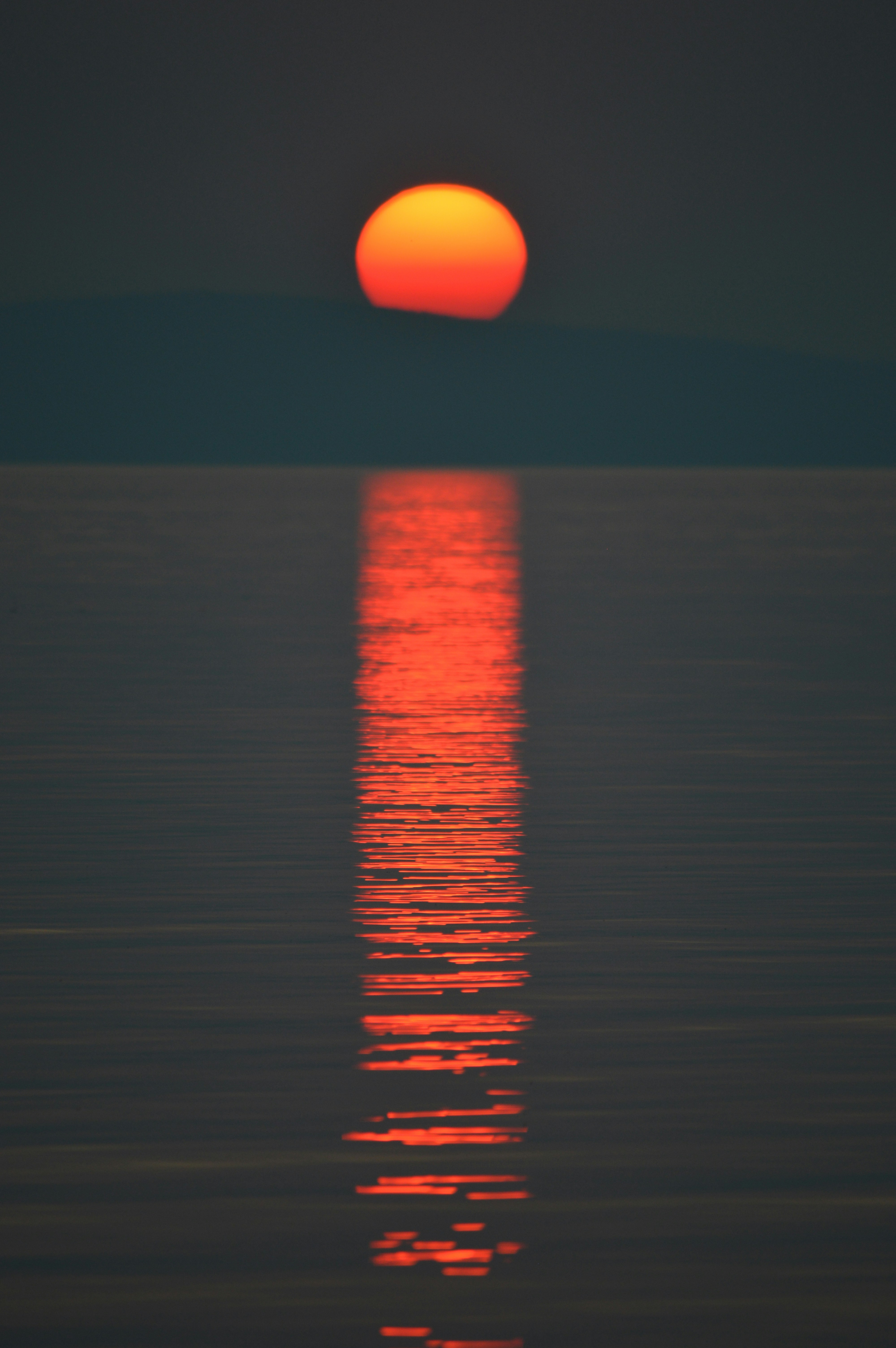 Vibrant orange sun setting over calm waters, casting a shimmering reflection that stretches towards the horizon.
