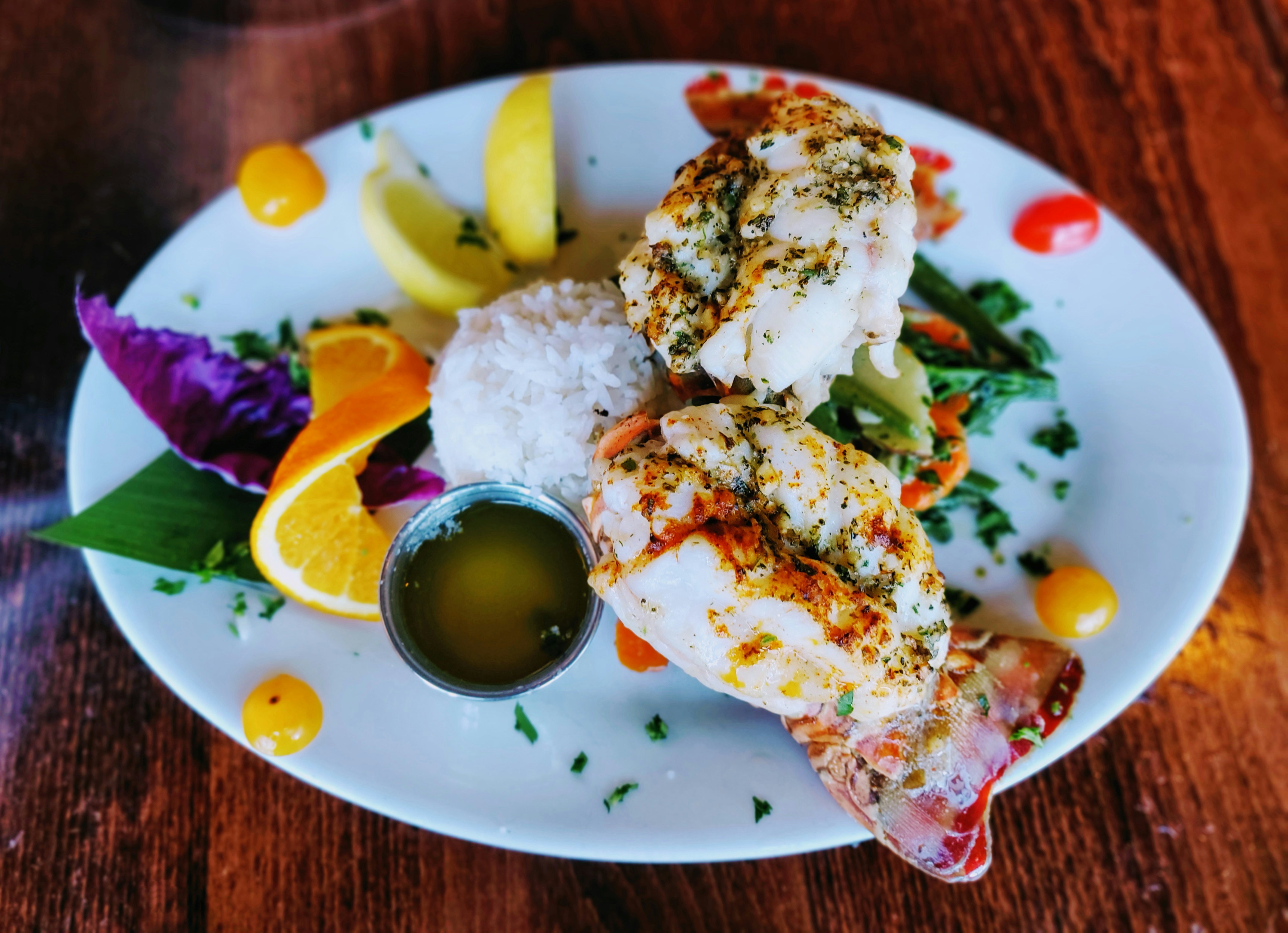 Grilled lobster tails garnished with herbs and served with rice, vegetables, and citrus slices on a white plate.