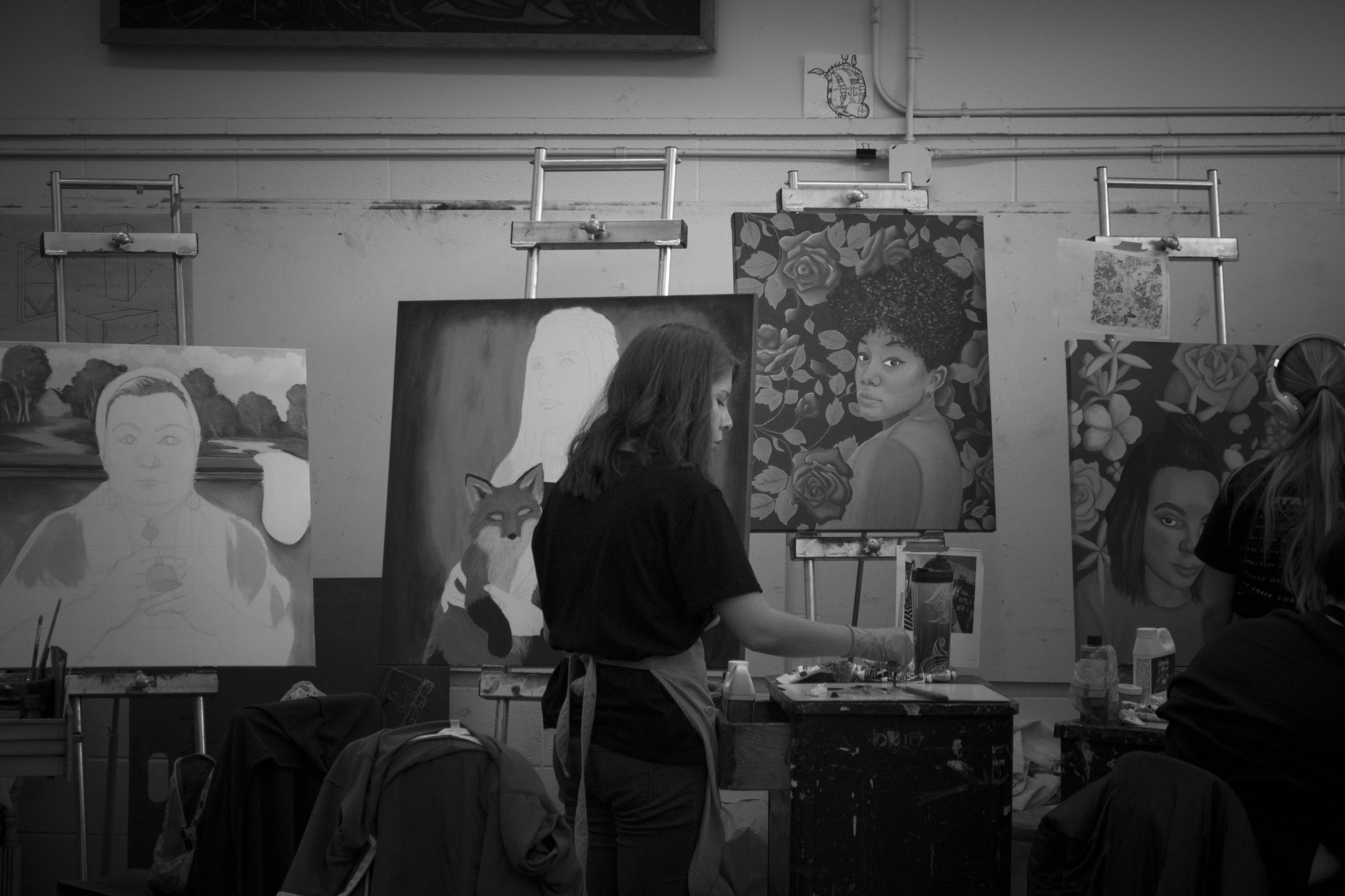 Artist working in a studio surrounded by portrait paintings on easels.