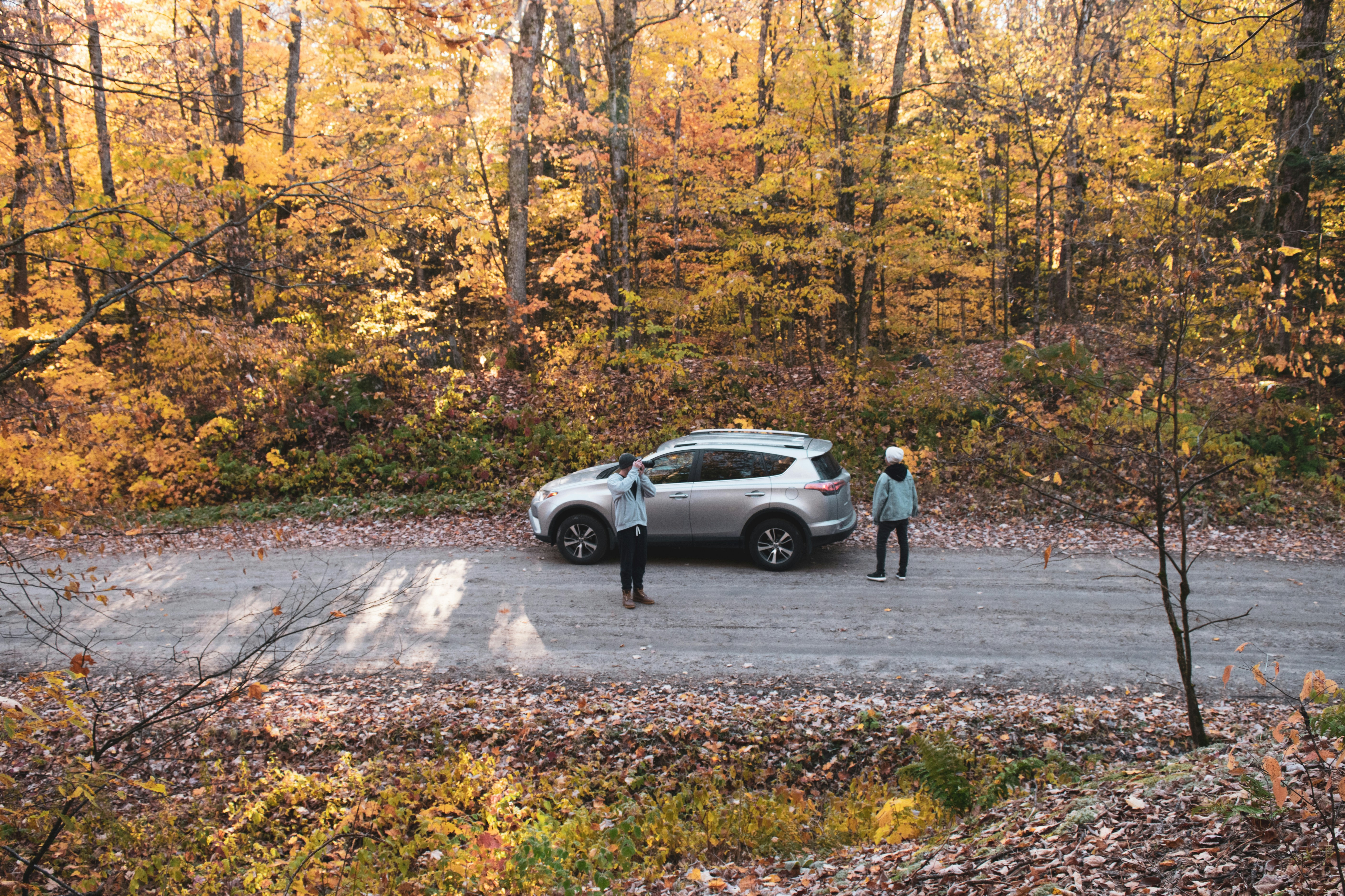 Two people stand by a silver car on a leaf-strewn road, surrounded by vibrant autumn foliage.