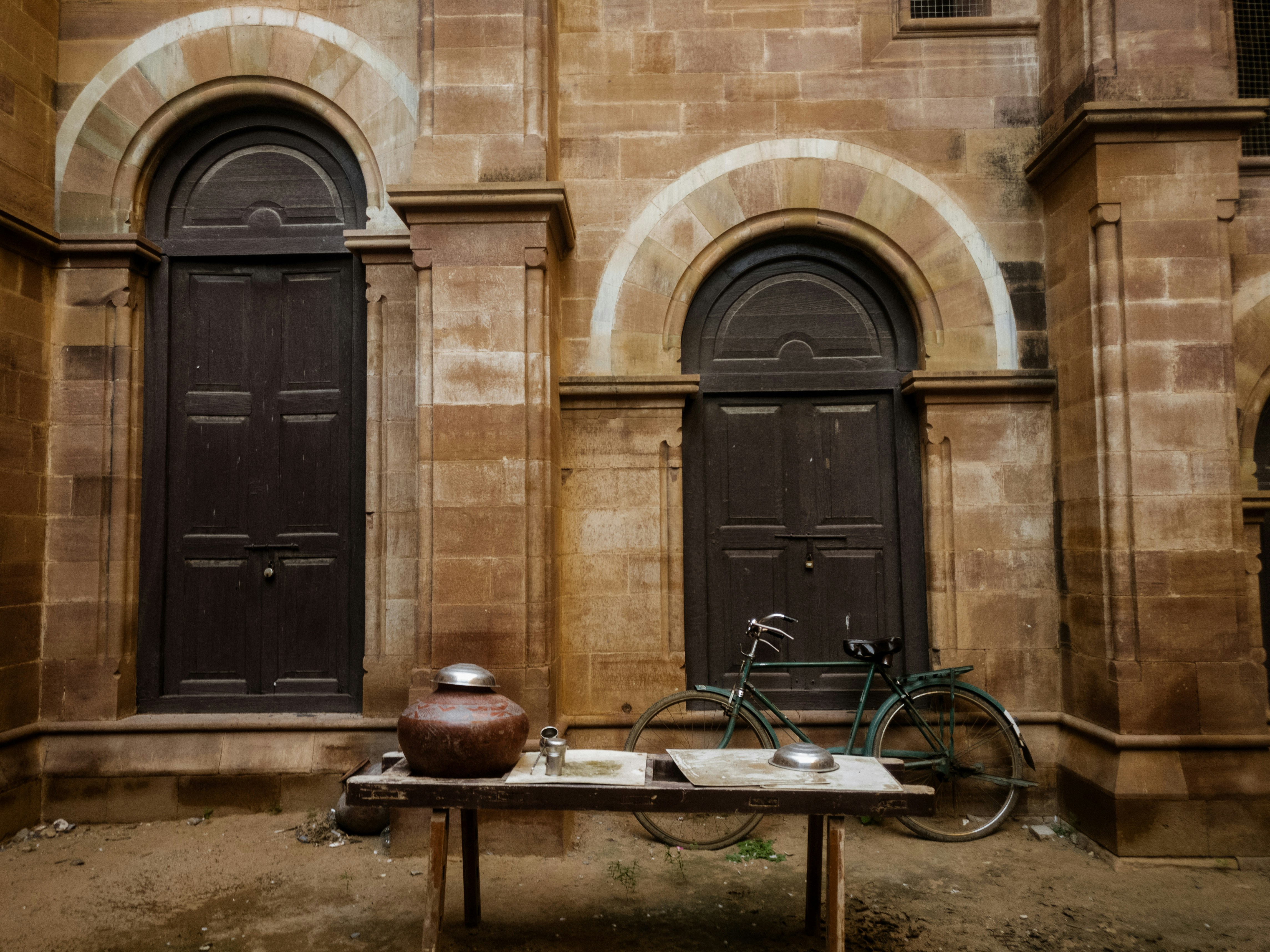 Weathered wooden table with clay pot and utensils against a historic brick building with arched doors.