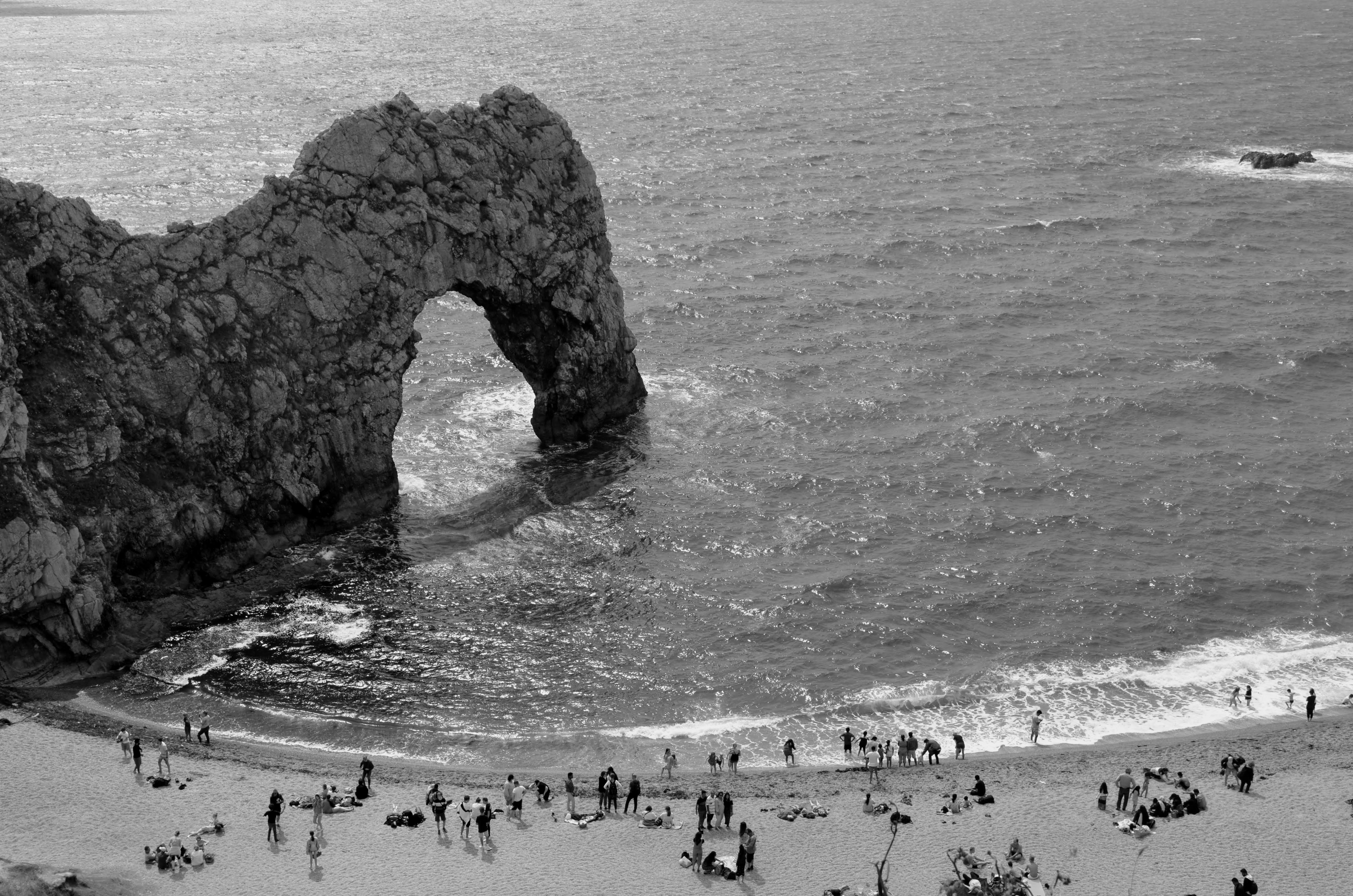 Black and white aerial view of a rocky archway at the seashore with people scattered along the beach.