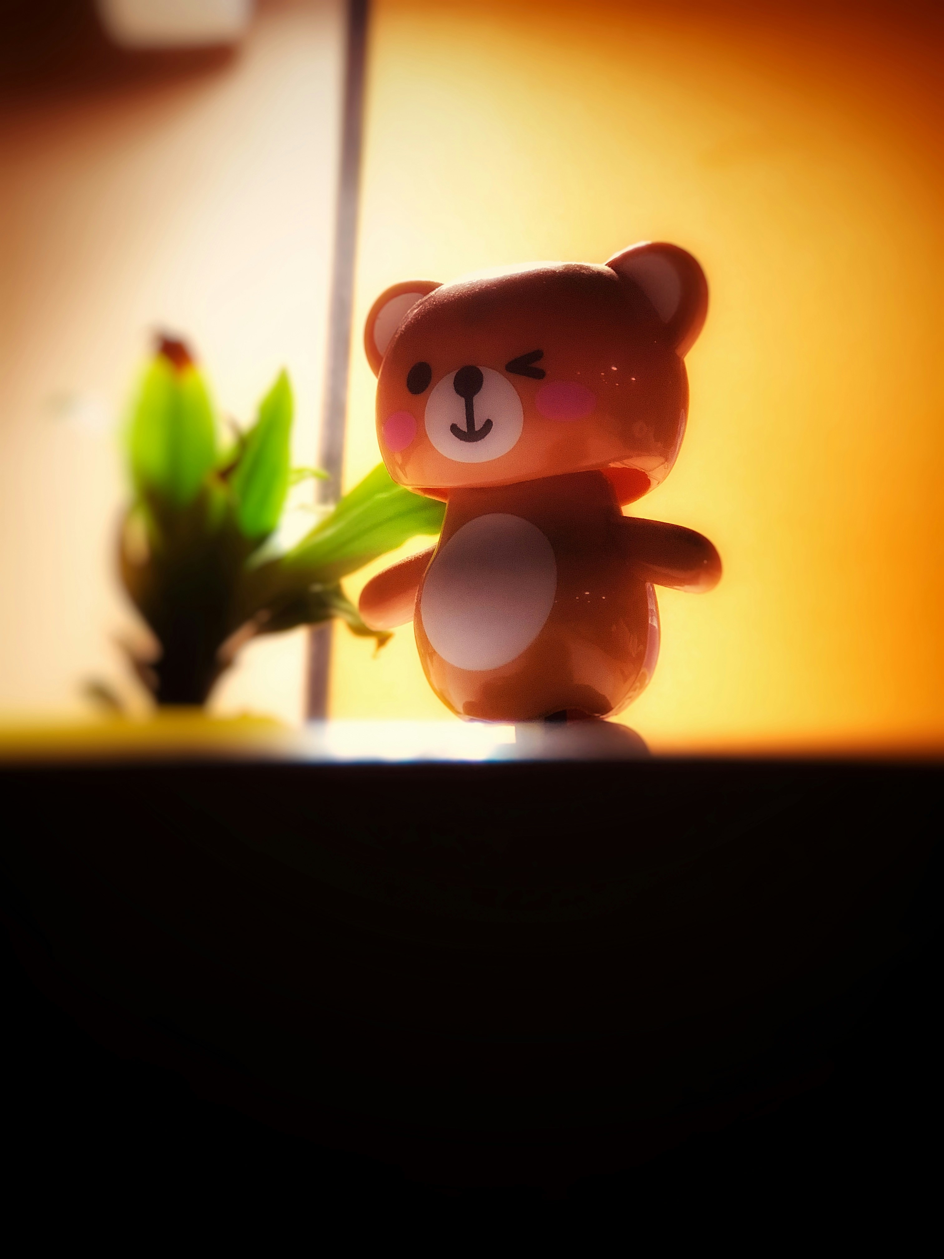 A playful bear figurine winks cheerfully against a softly lit backdrop, with a hint of greenery nearby.