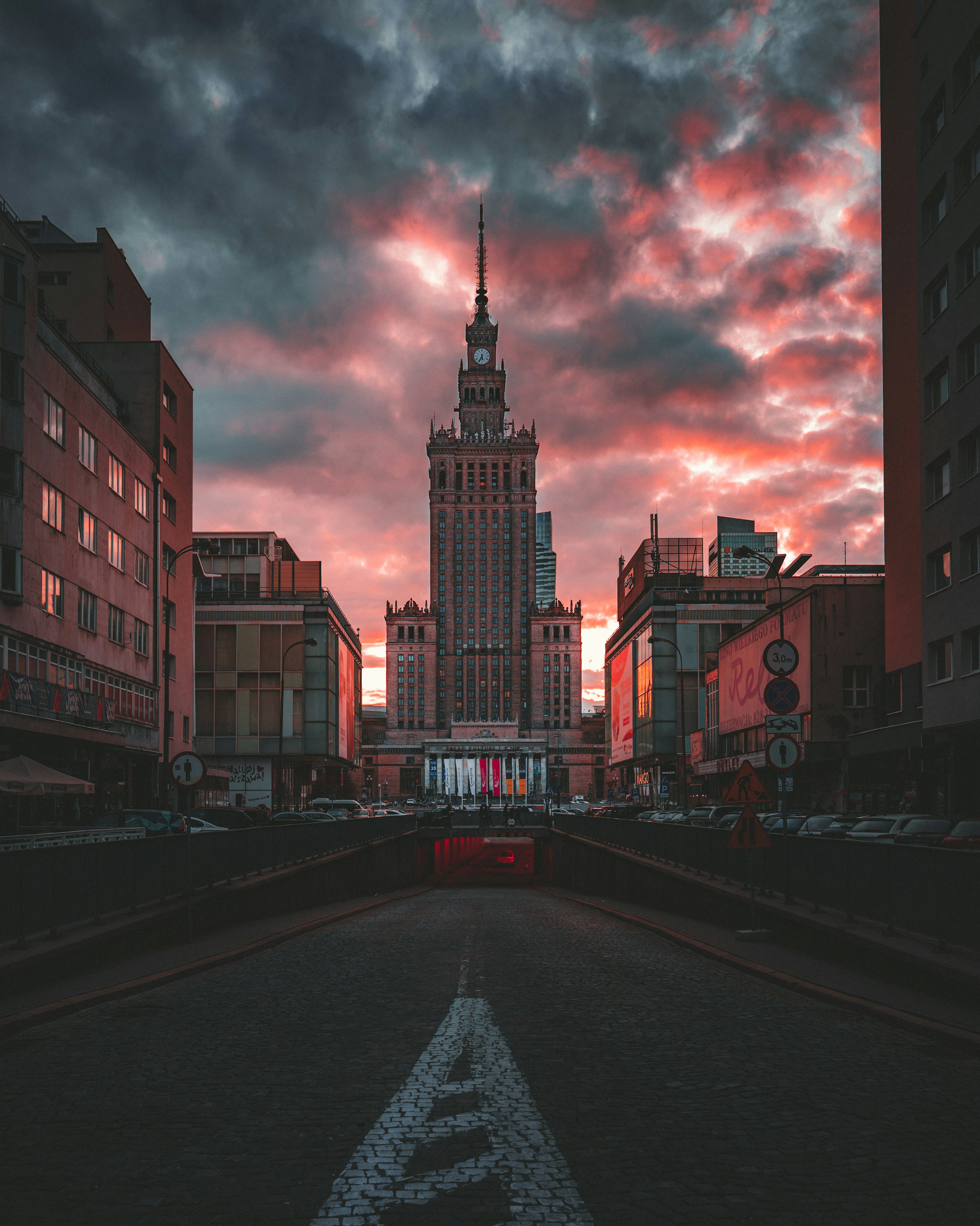The iconic Palace of Culture and Science stands tall amidst a dramatic sunset, framed by modern city buildings. A hint of urban life is visible with cars lining the streets below.